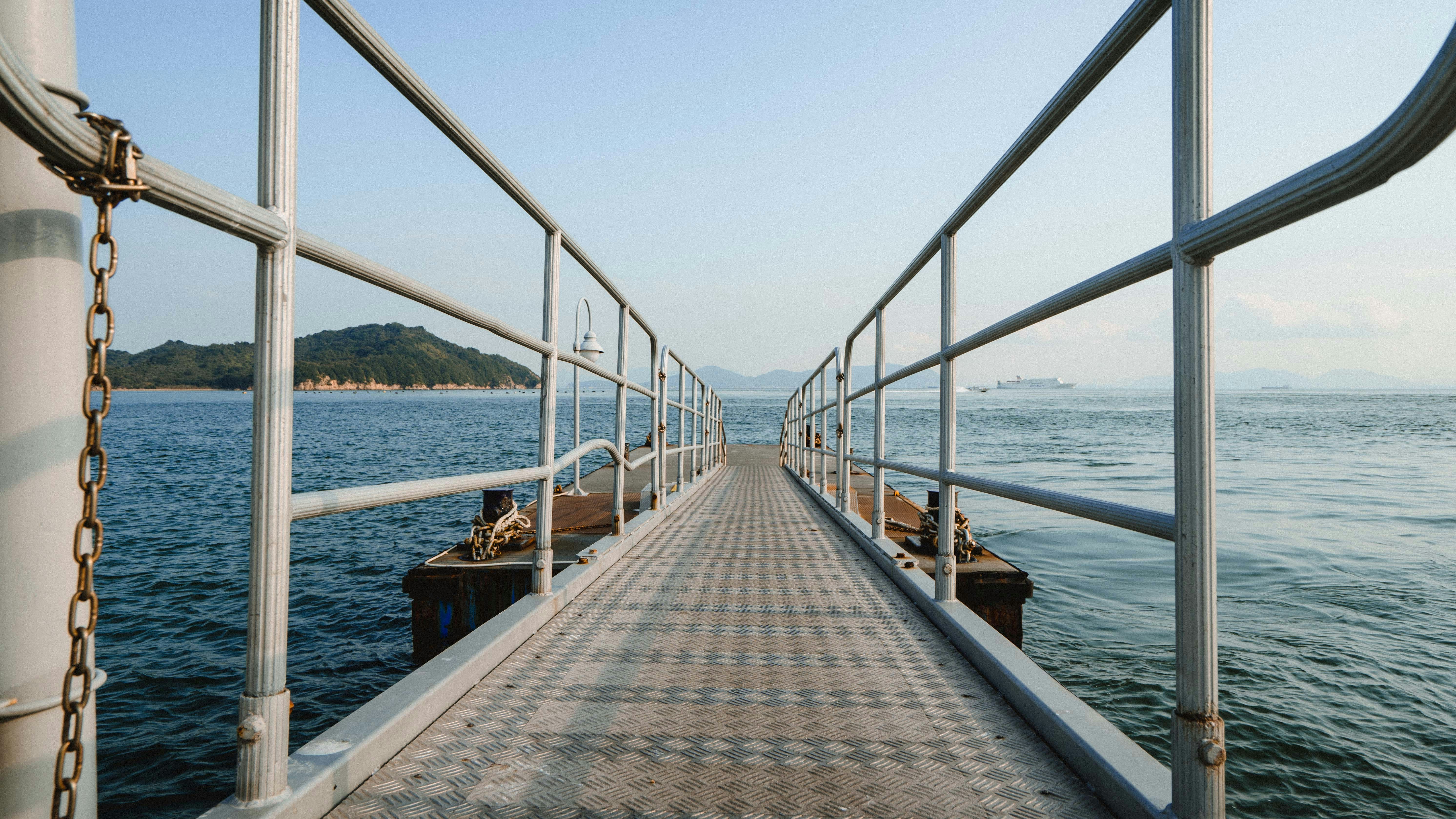 Metal pier extends into tranquil ocean with distant islands and ships.