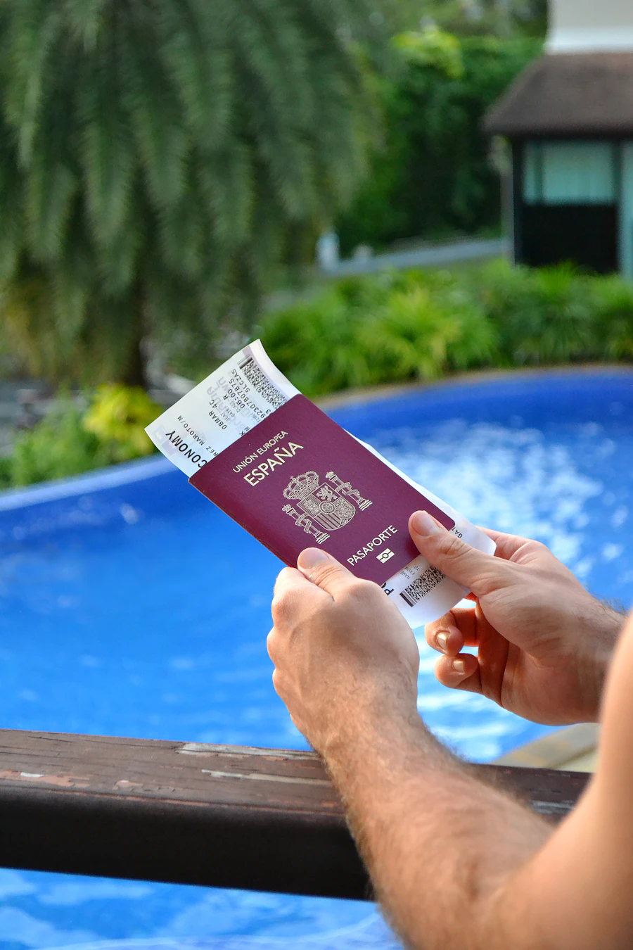 Hands holding a Spanish passport with a boarding pass tucked inside, set against a softly blurred blue pool and tropical greenery background.