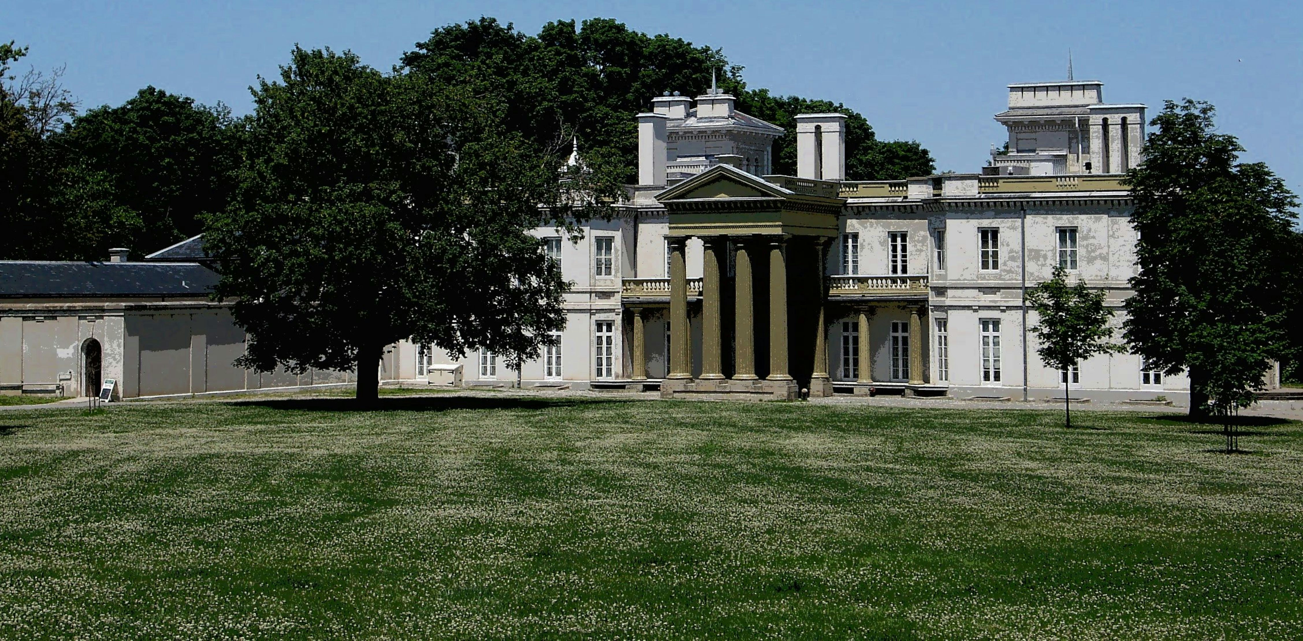 Large mansion with prominent columns stands in a lush green field surrounded by trees.