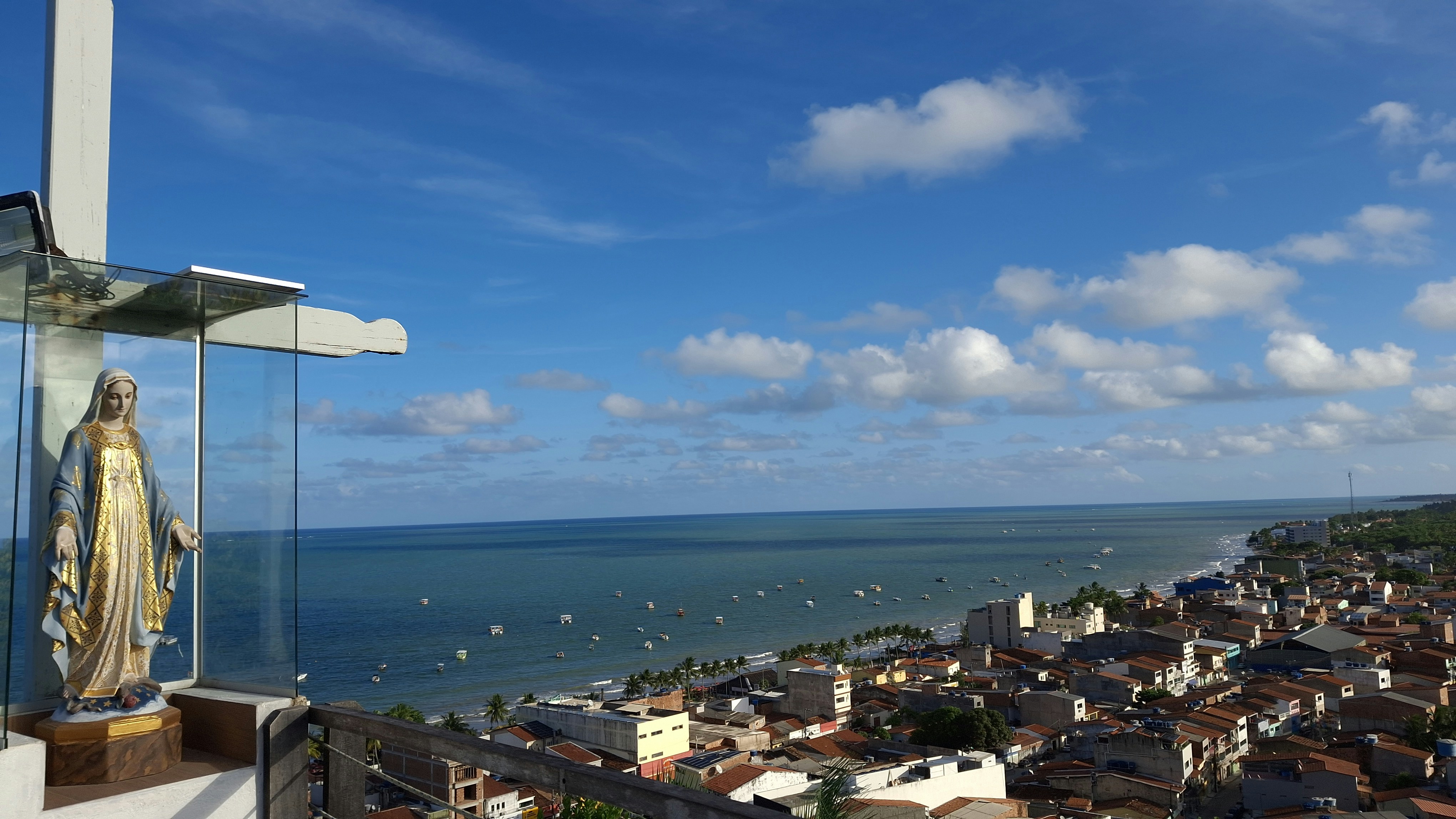 Statue of a figure in a glass case overlooking a coastal town with a vast ocean stretching to the horizon under a clear blue sky.
