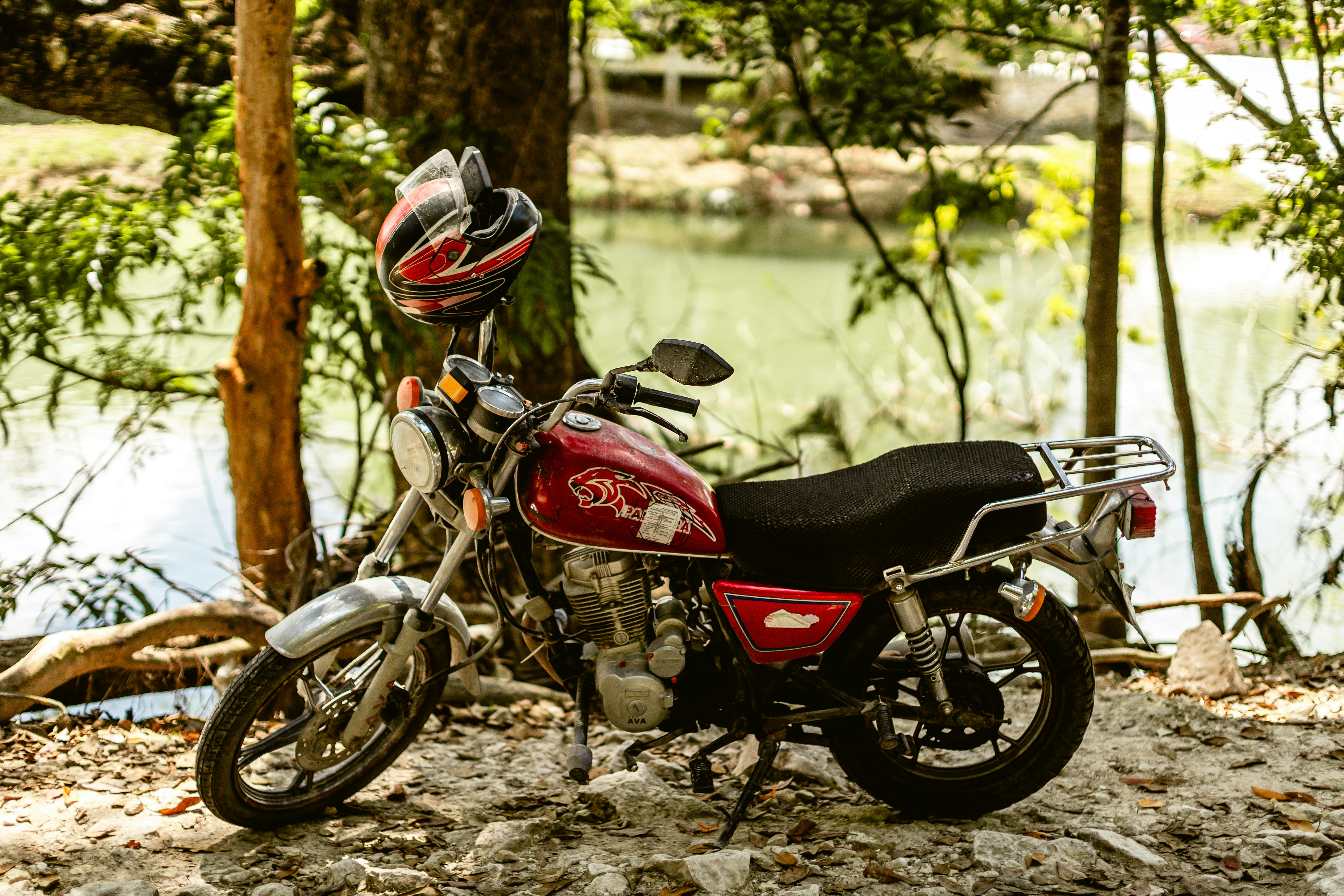 Red motorcycle parked on rocky ground near a serene riverbank surrounded by trees.