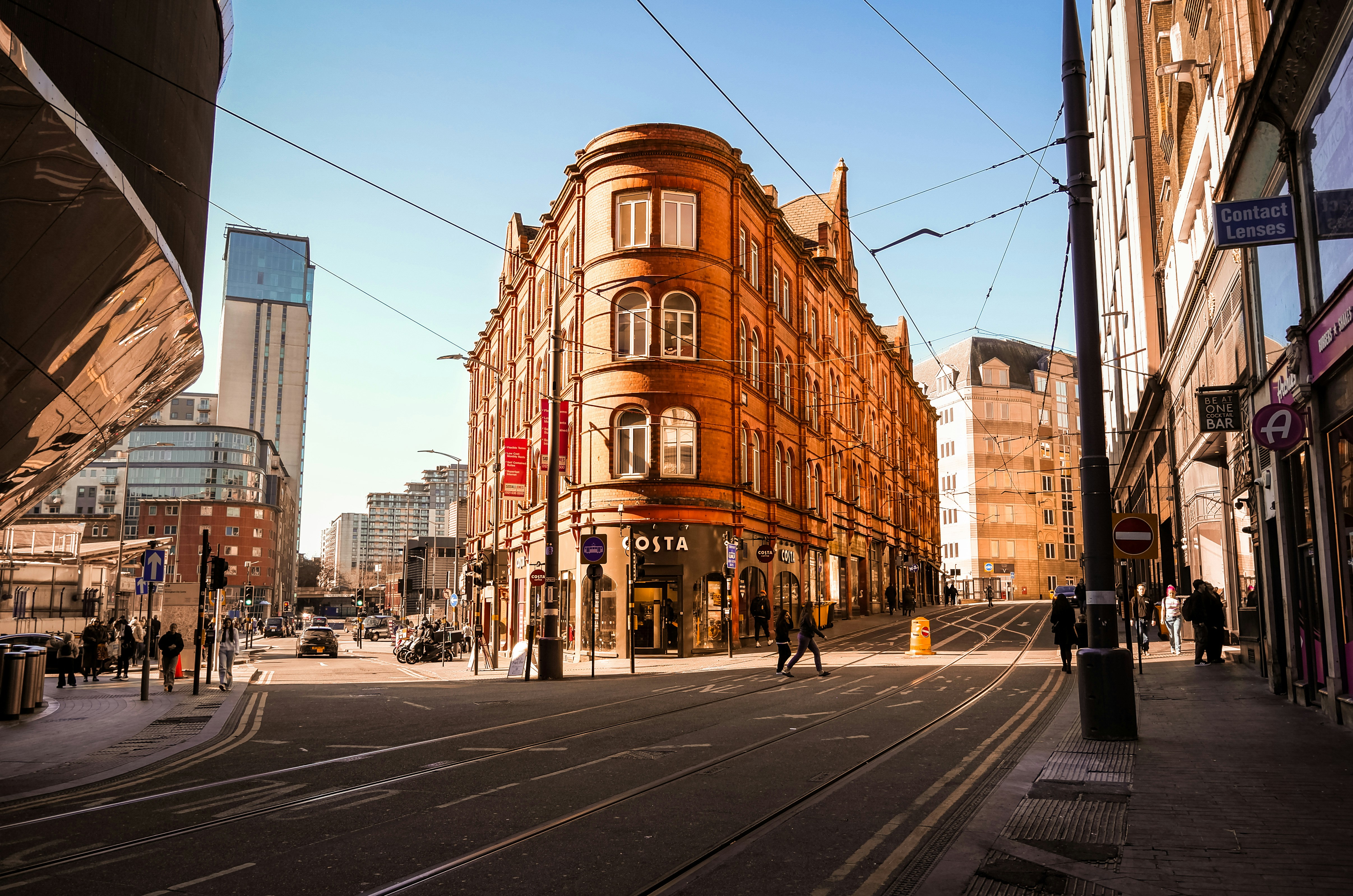 Sunlit city street centered on a curved red-brick corner building, with overhead tram wires, pedestrians, and rails.
