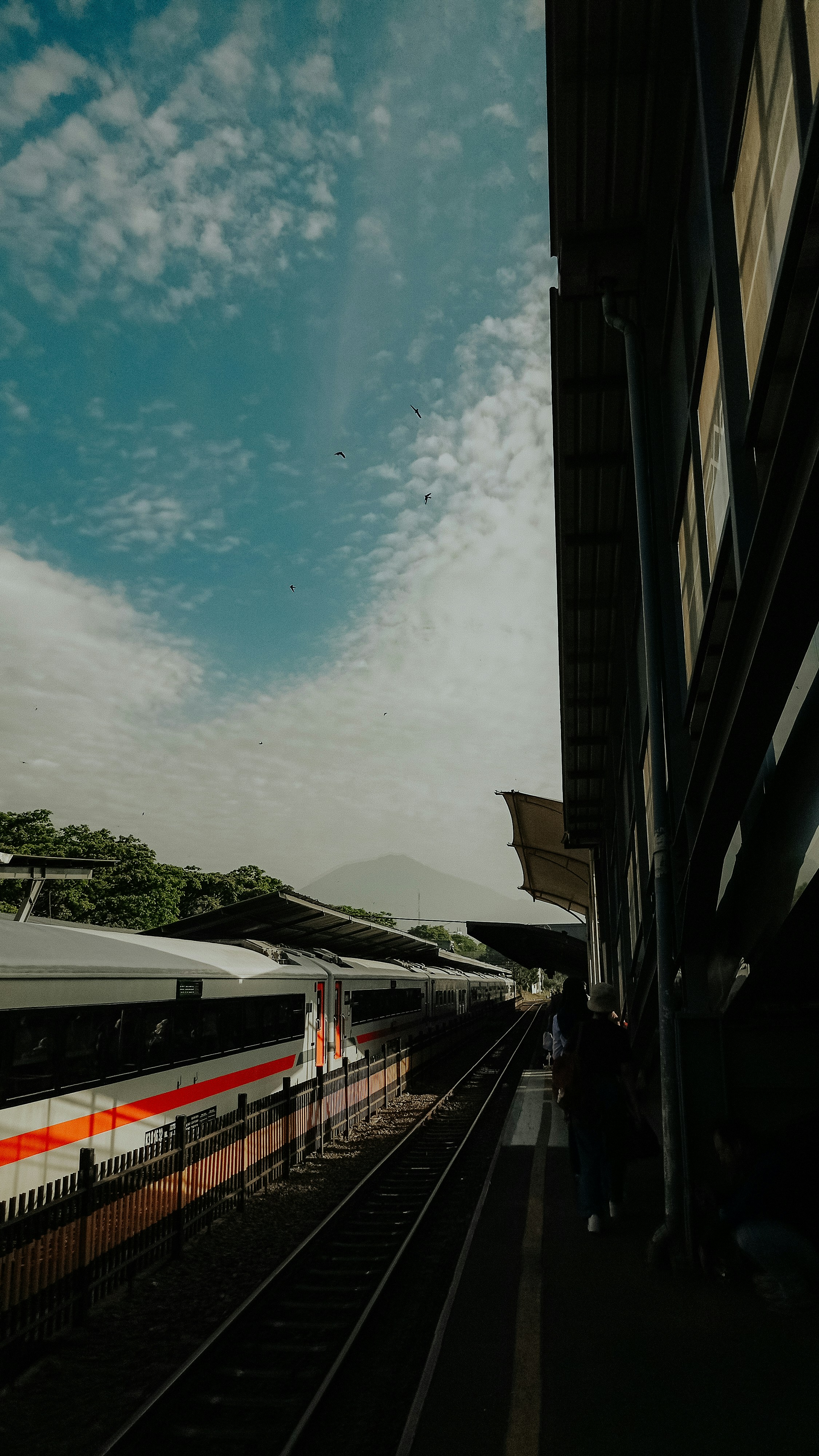 A sleek train awaits at a station under a vast sky, with mountains in the background and passengers nearby.