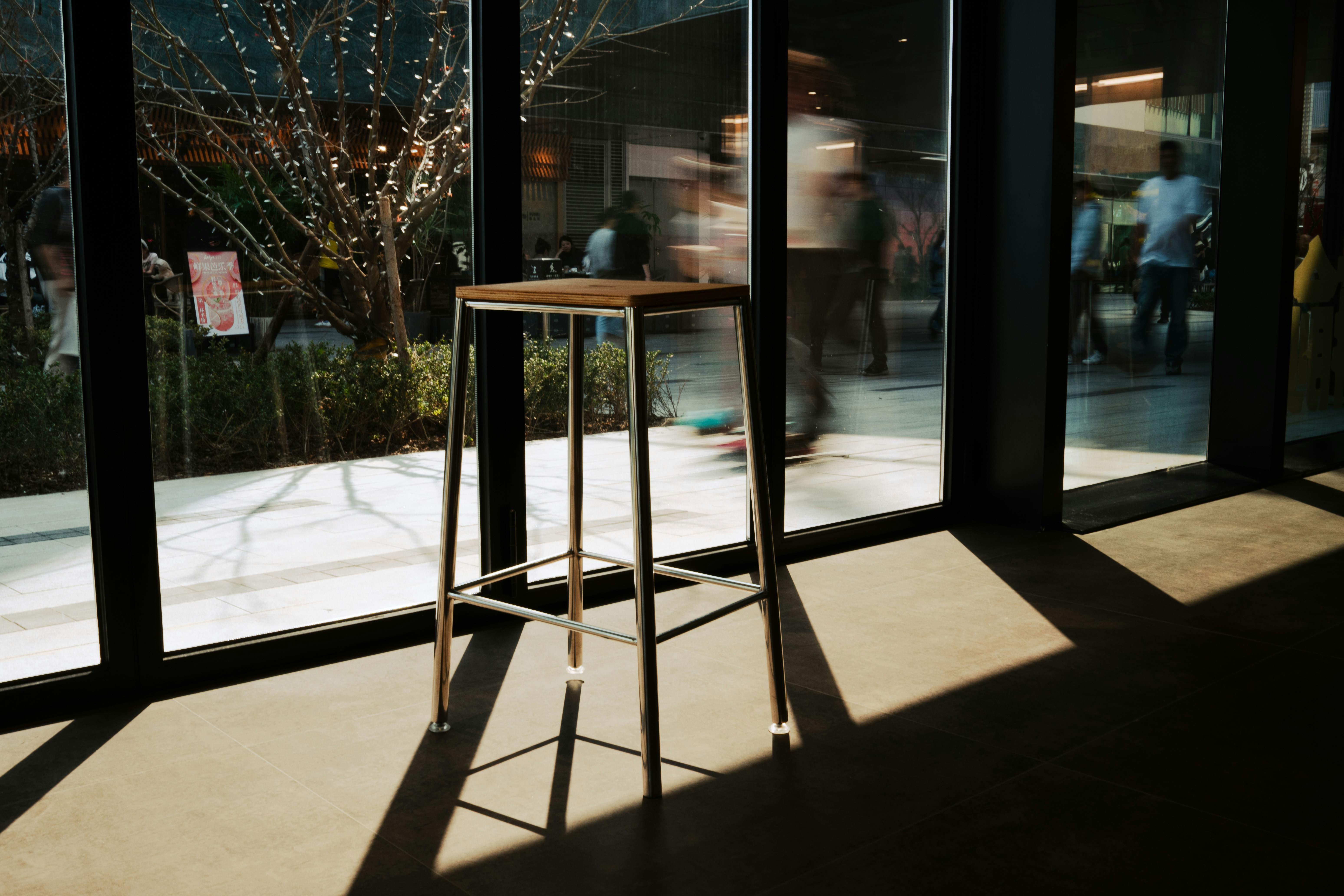 A solitary stool in soft sunlight casts a shadow against a bustling urban backdrop through a window.