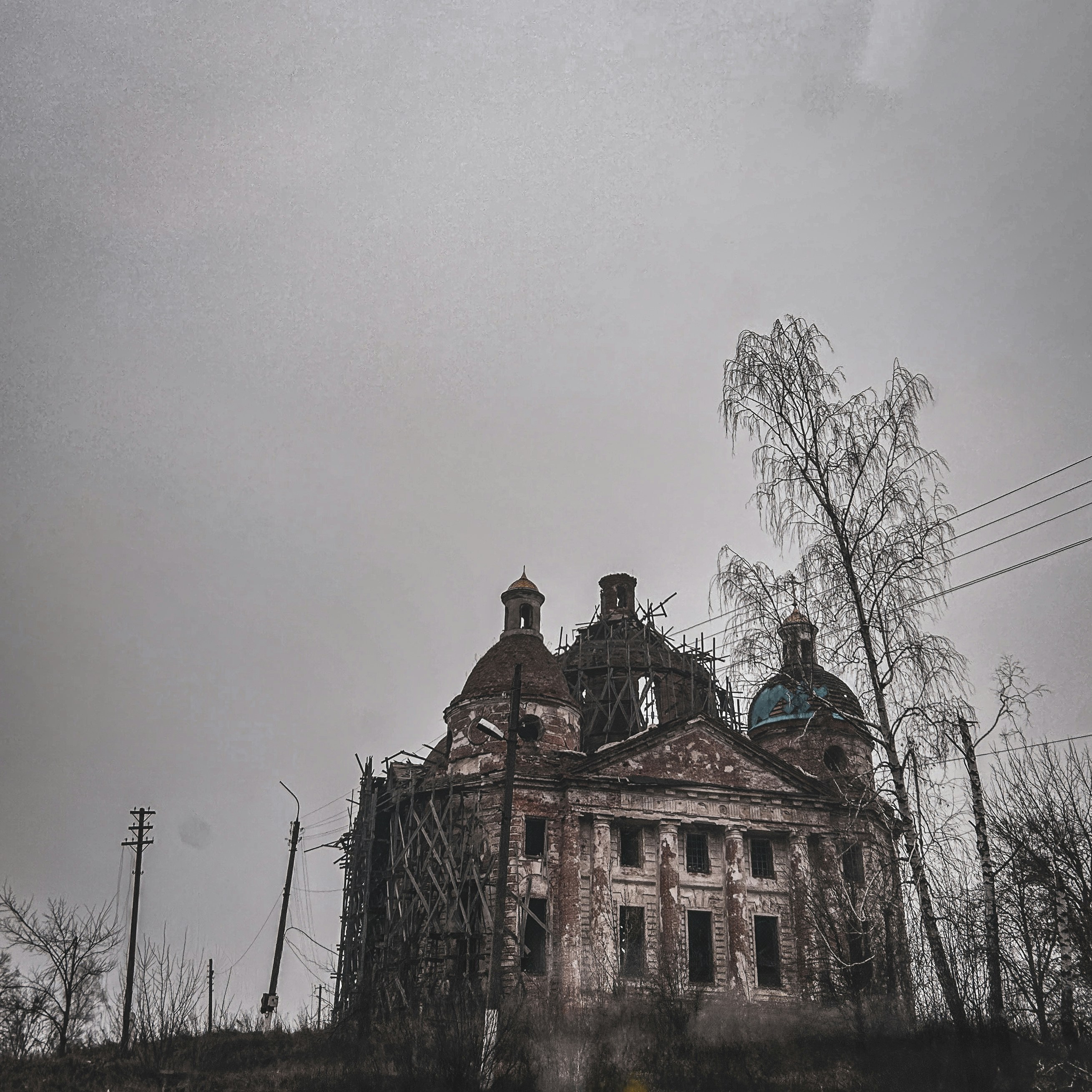 Ruined church with exposed scaffolding and bare trees beneath a moody, overcast sky.