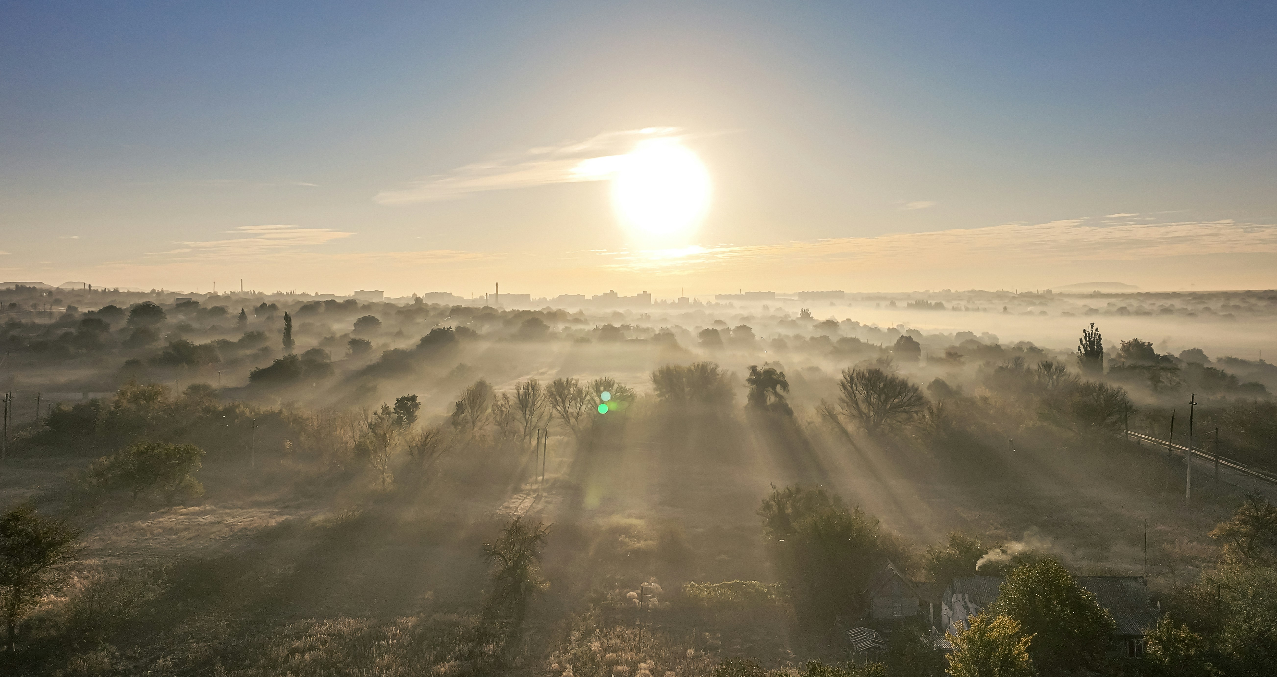 Sunrise illuminates a fog-laden landscape with rays piercing through trees and mist.