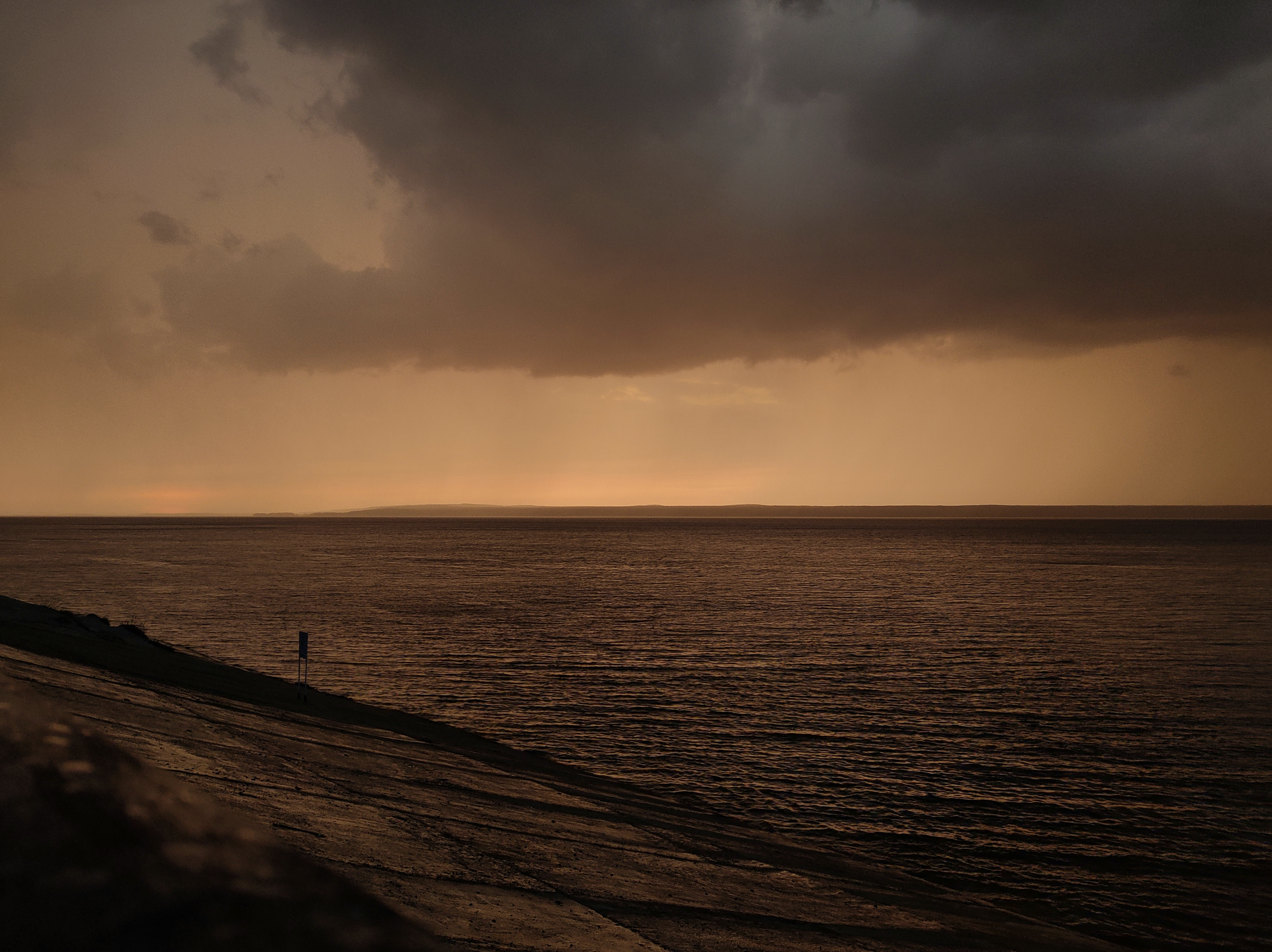 Dark clouds loom over a calm river with a golden sunset in the distance.