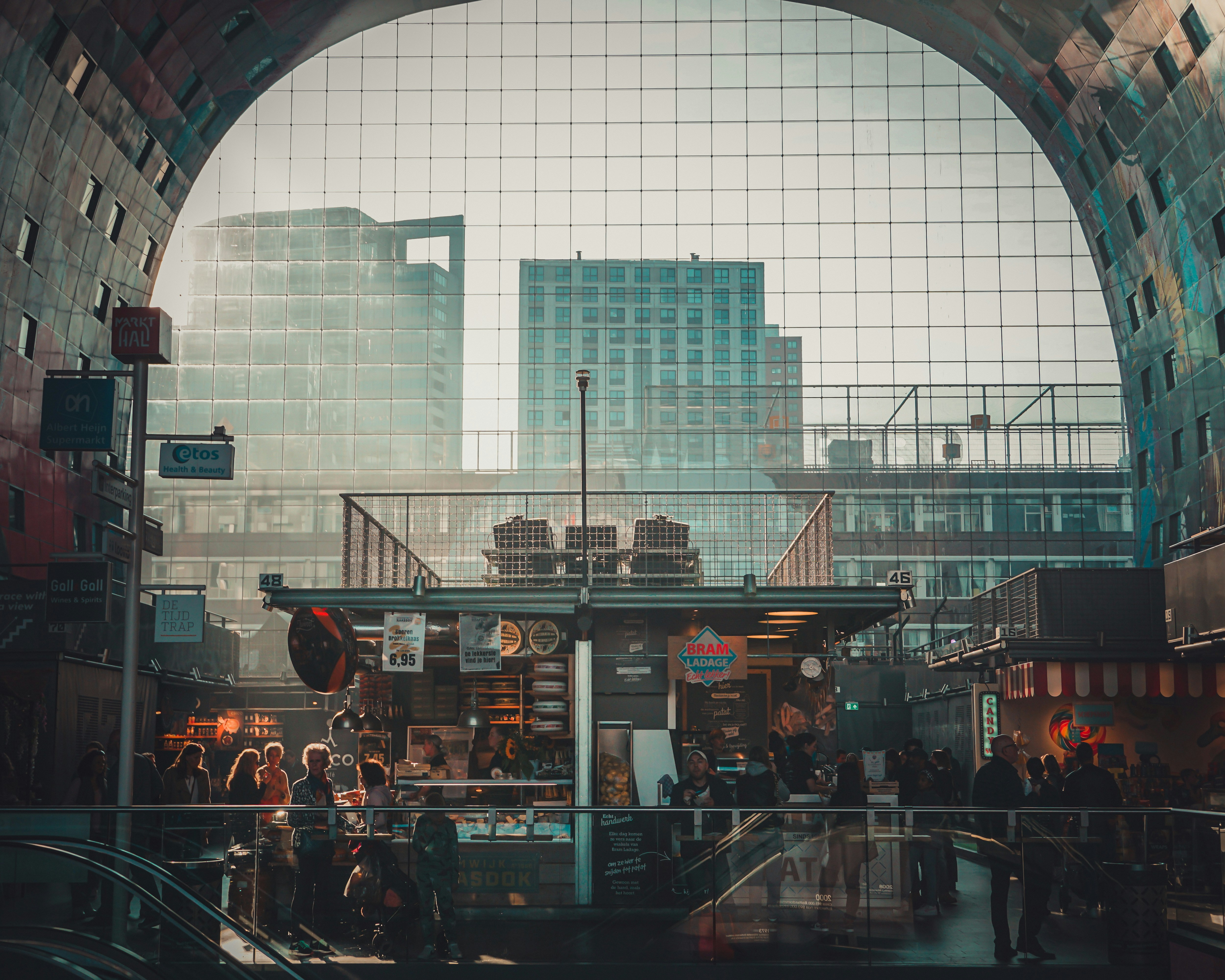 Shoppers navigate a bustling market beneath a vast arched glass ceiling, with sunlight streaming through modern architecture.
