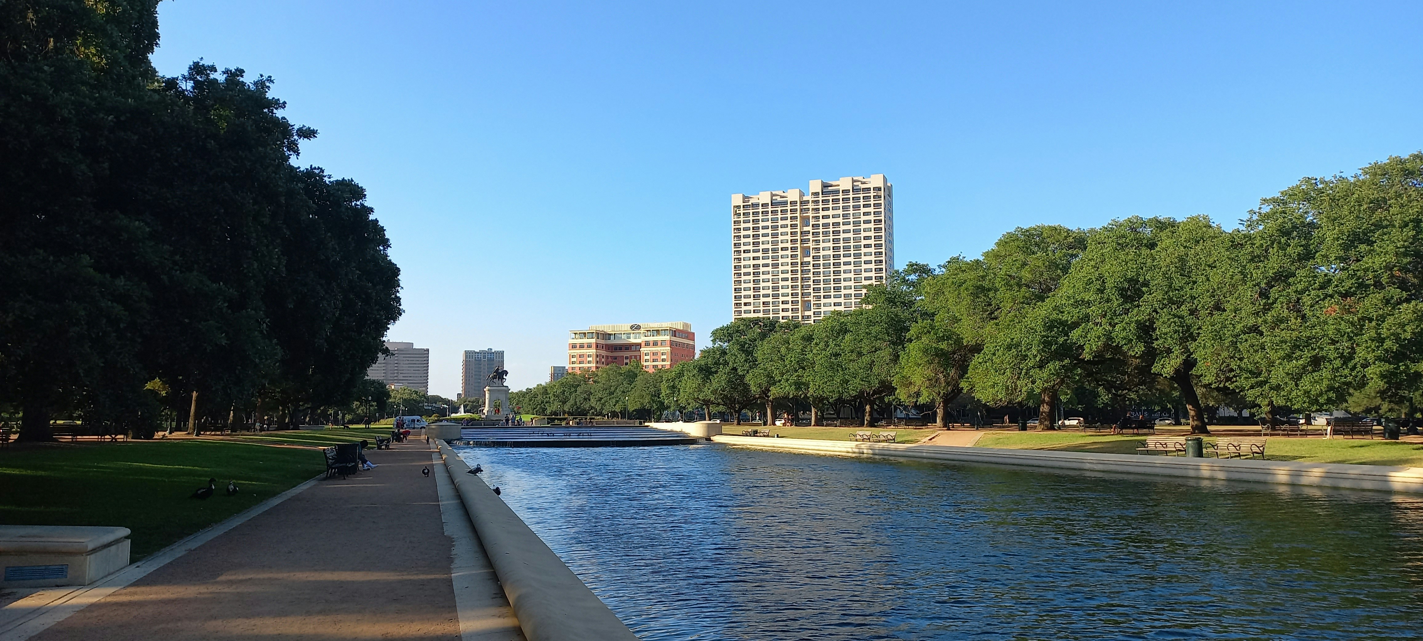 City park canal photograph with a sunlit waterway flanked by tree-lined banks and a distant high-rise skyline.