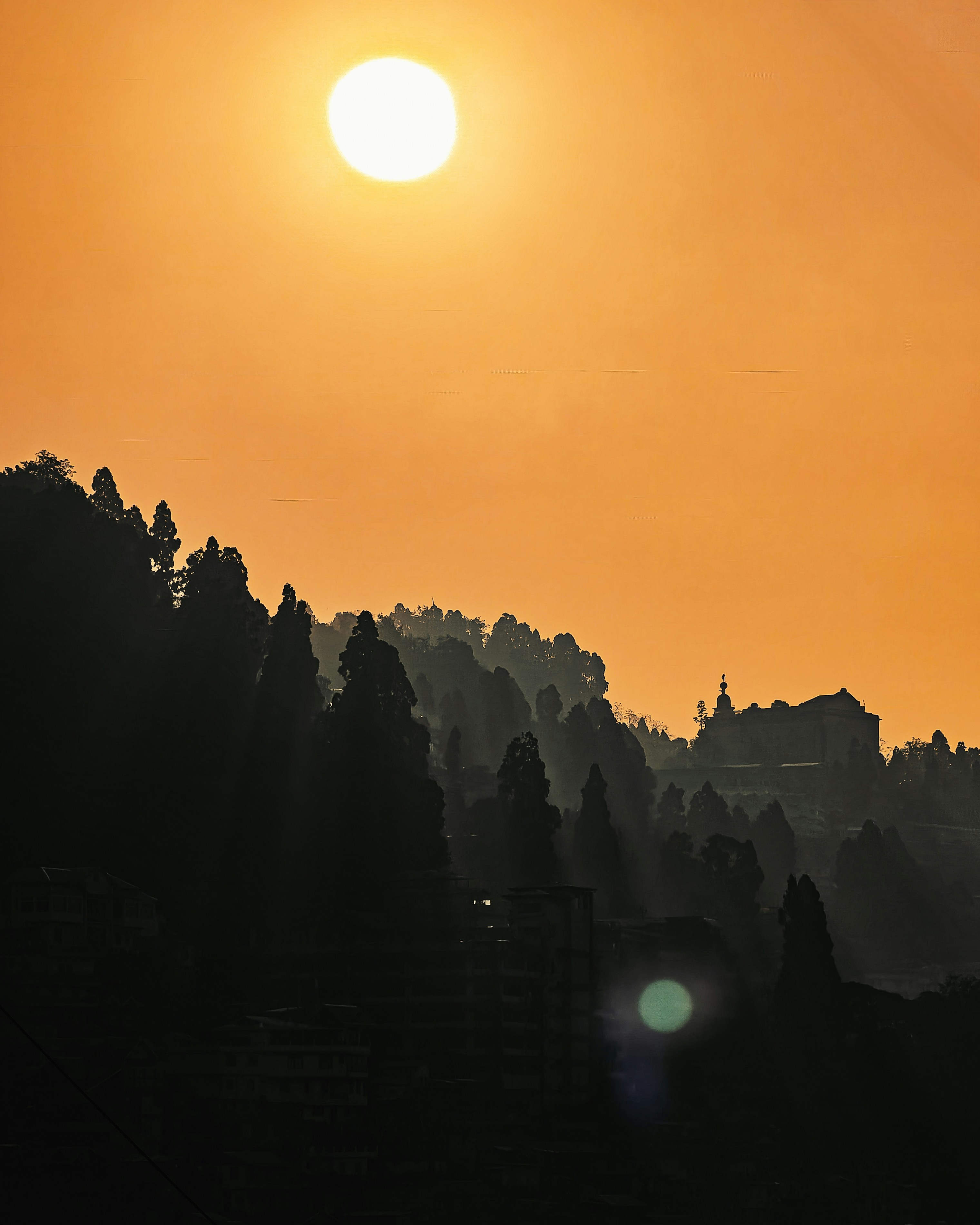 Silhouetted trees and a distant building under a vibrant orange sunset sky.