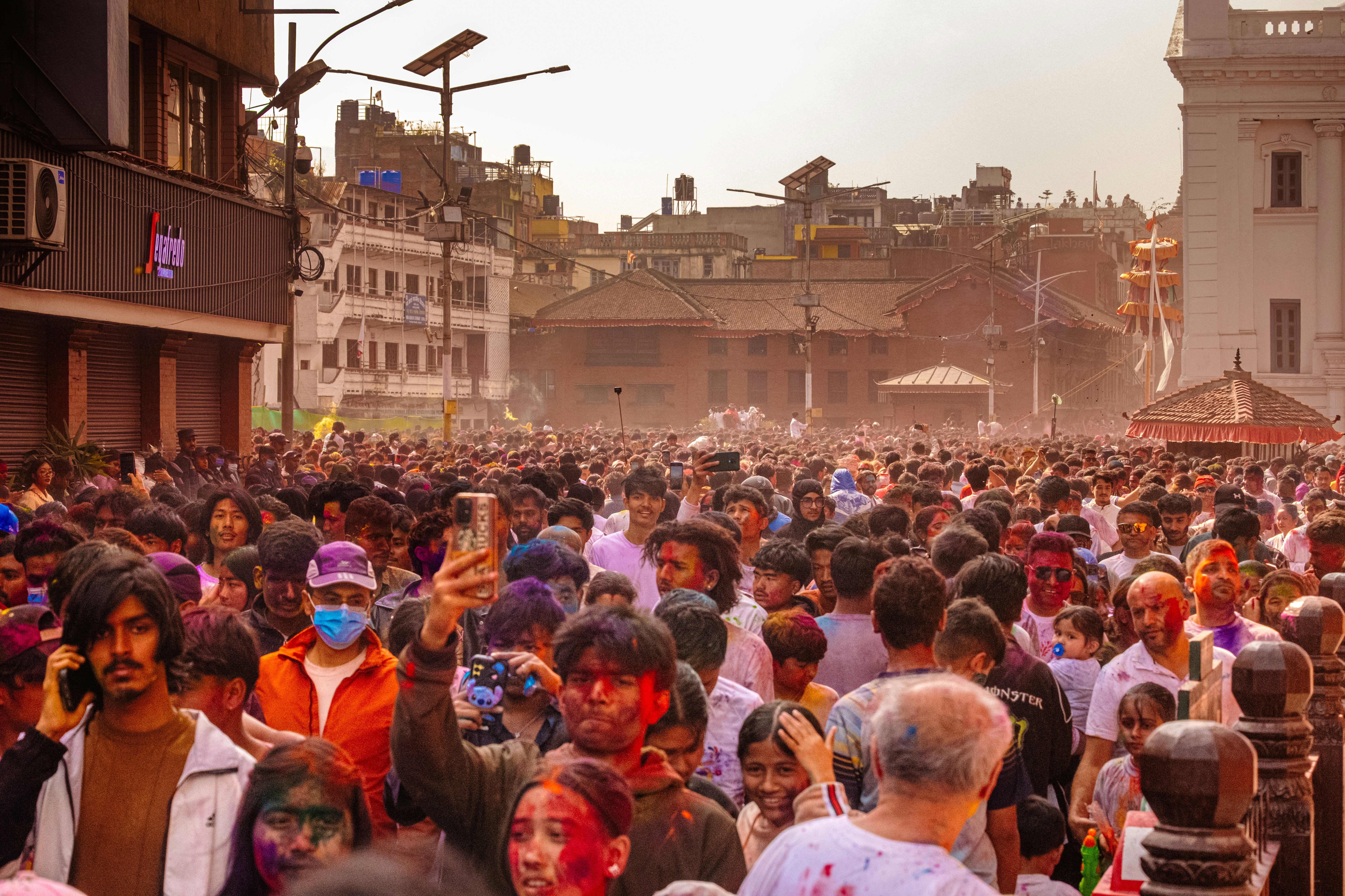 Crowd celebrating a vibrant festival in a city square, with buildings in the background and colorful powders in the air.