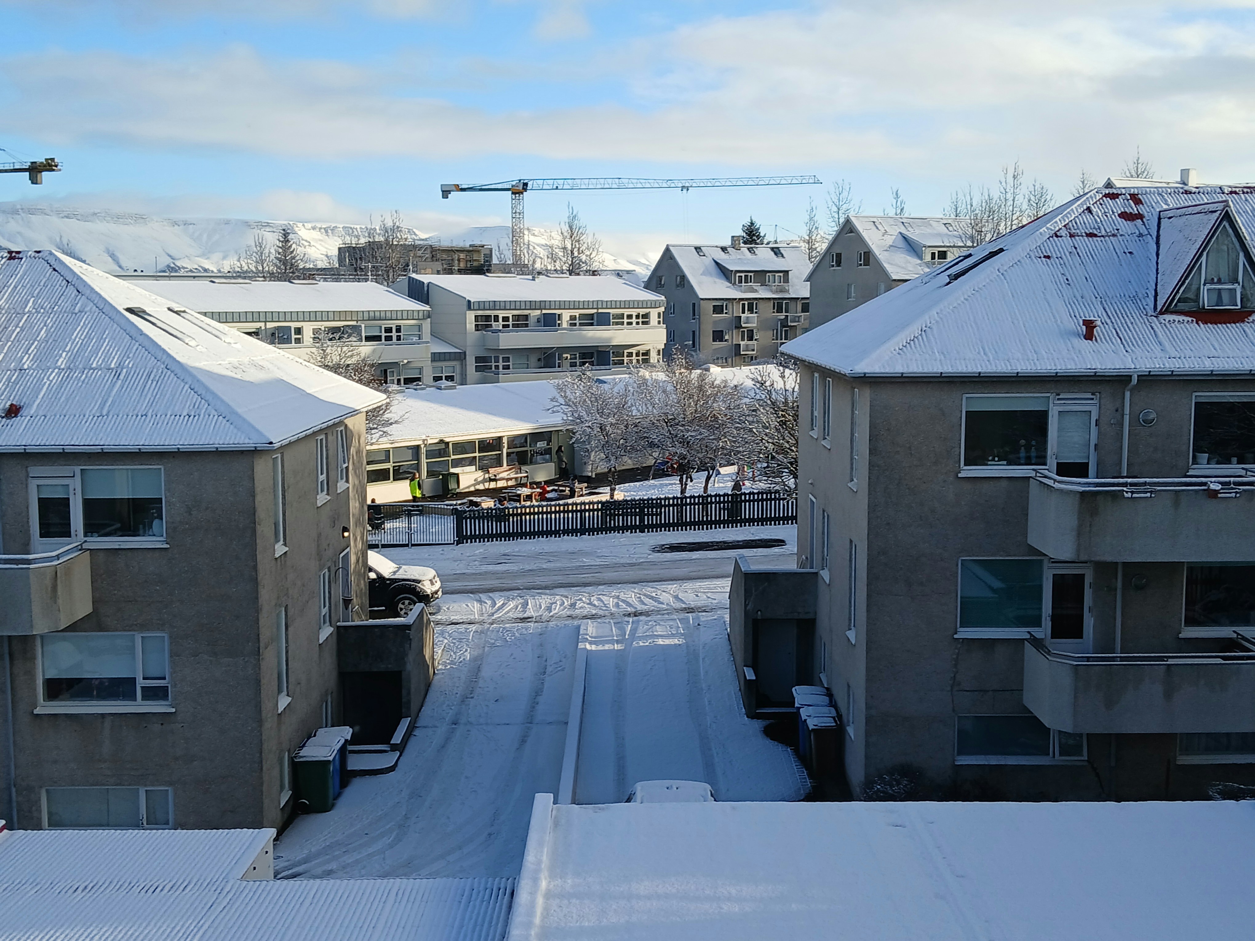 Snow-covered rooftops and streets with distant cranes under a bright winter sky.
