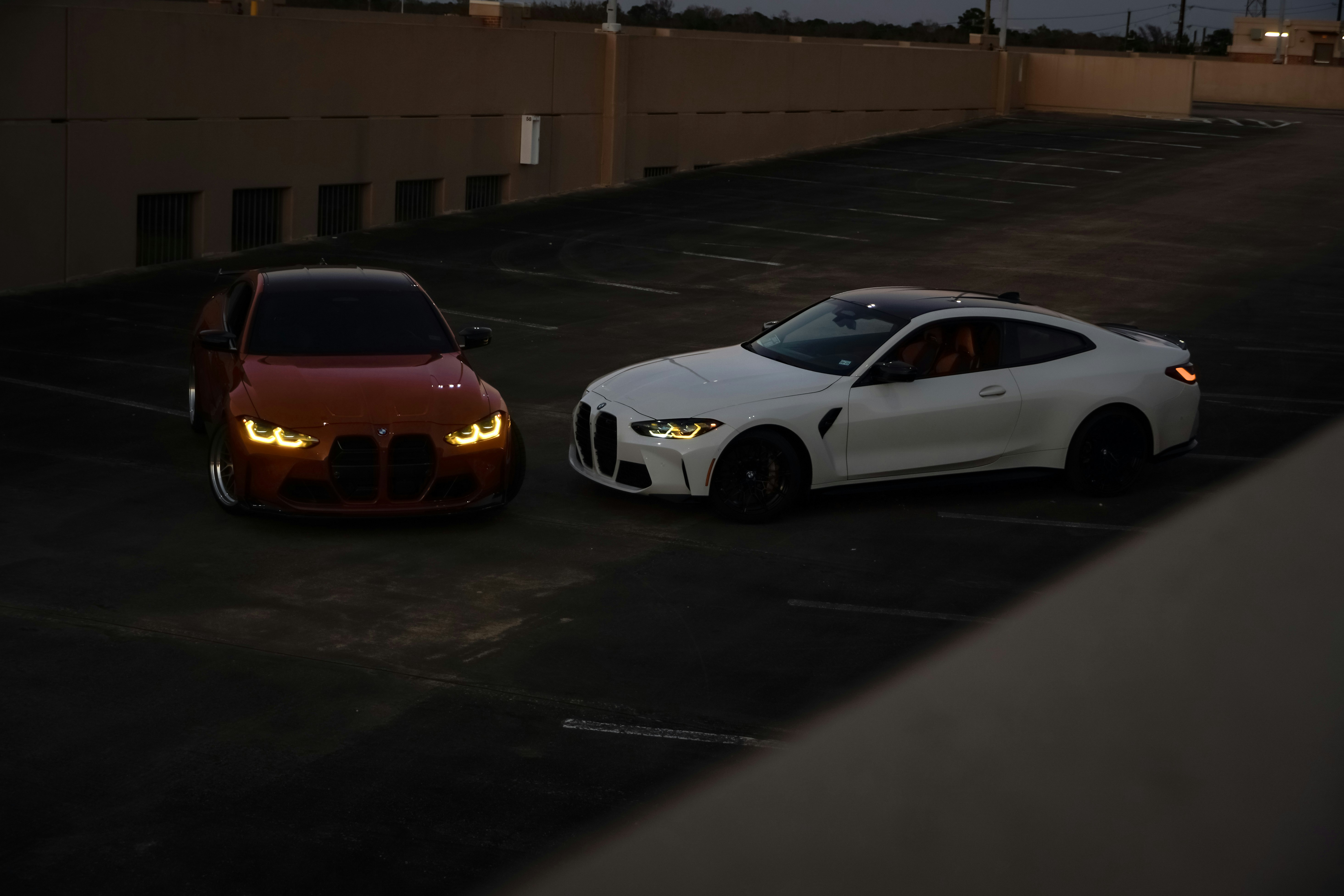 Two cars parked in a dimly lit urban parking lot, headlights glowing in the dusk.