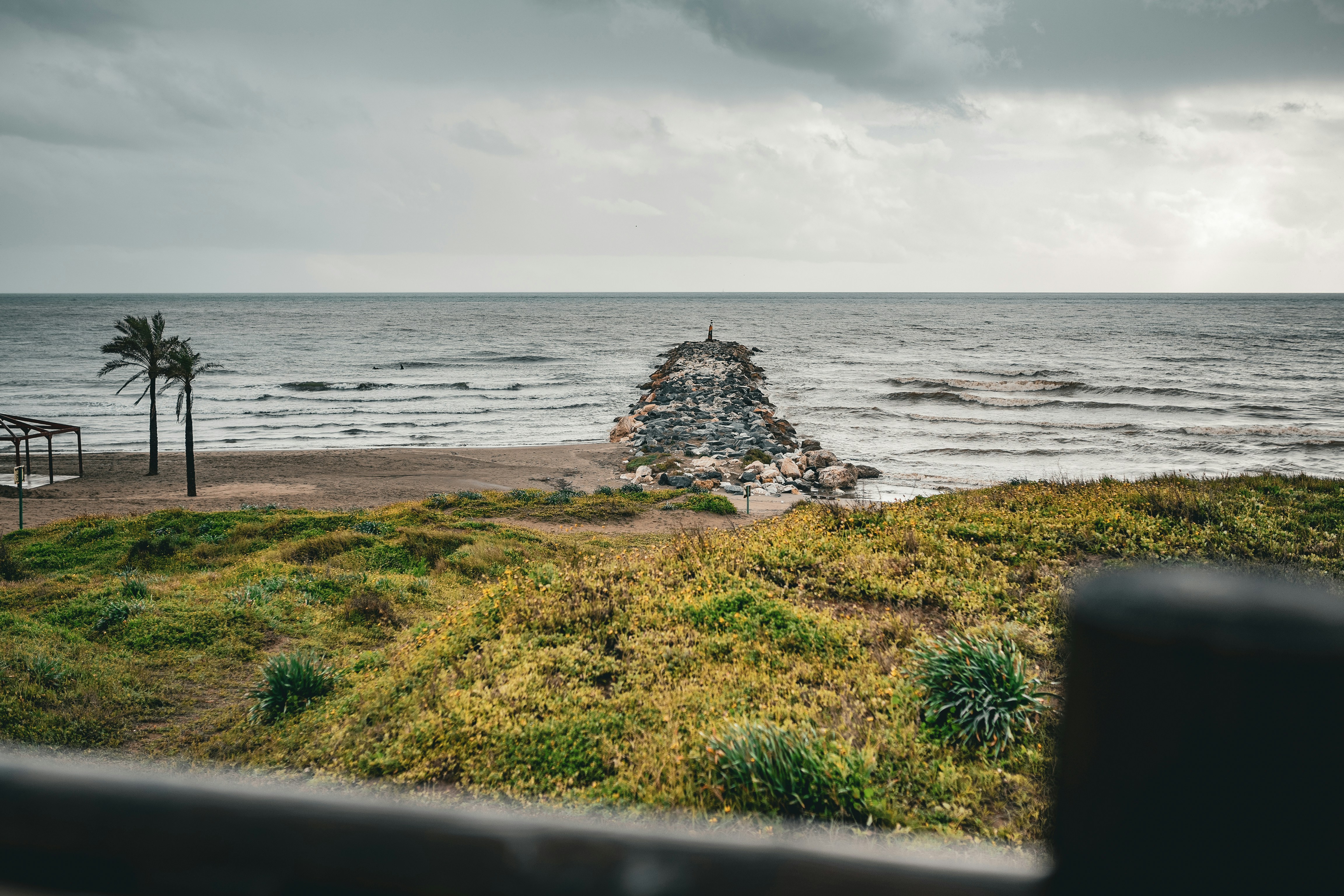 Stone pier extends into the sea beneath a cloudy sky, flanked by sparse palm trees and lush green foreground.