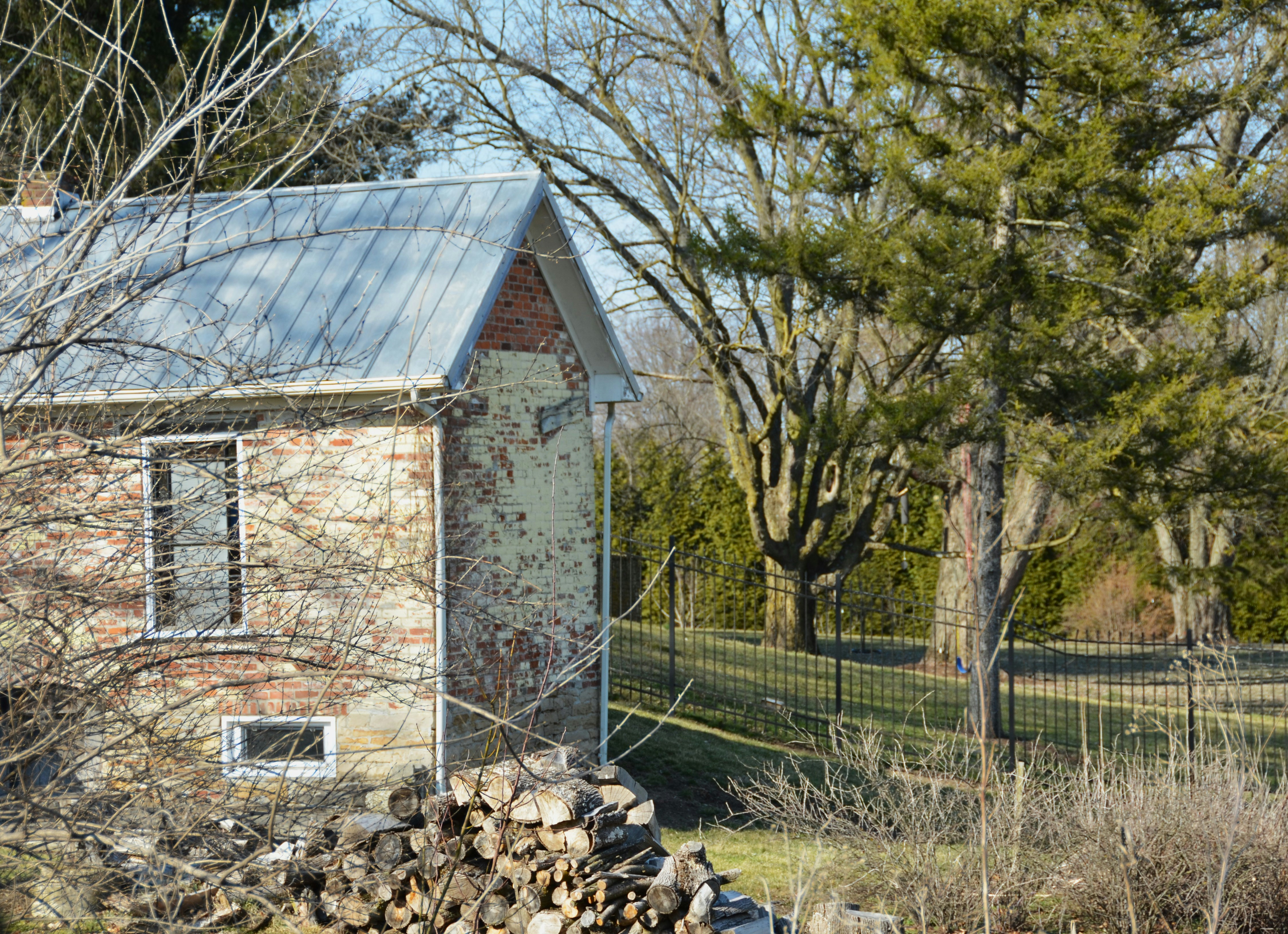 Weathered brick house with a tin roof stands near leafless trees and a stack of firewood.