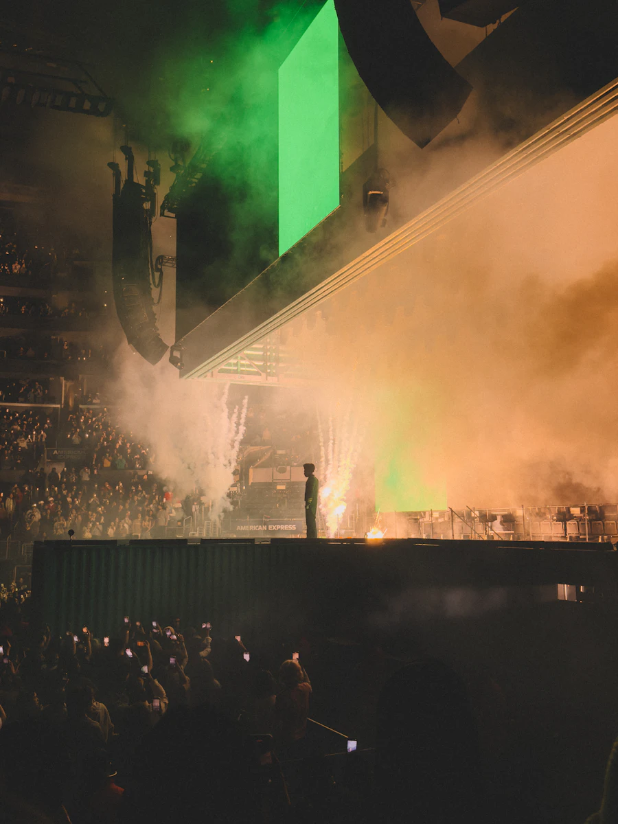 A lone performer stands on a raised stage amid heavy smoke, bright green screens, and warm orange light in a packed arena. The crowd fills the lower half of the scene.