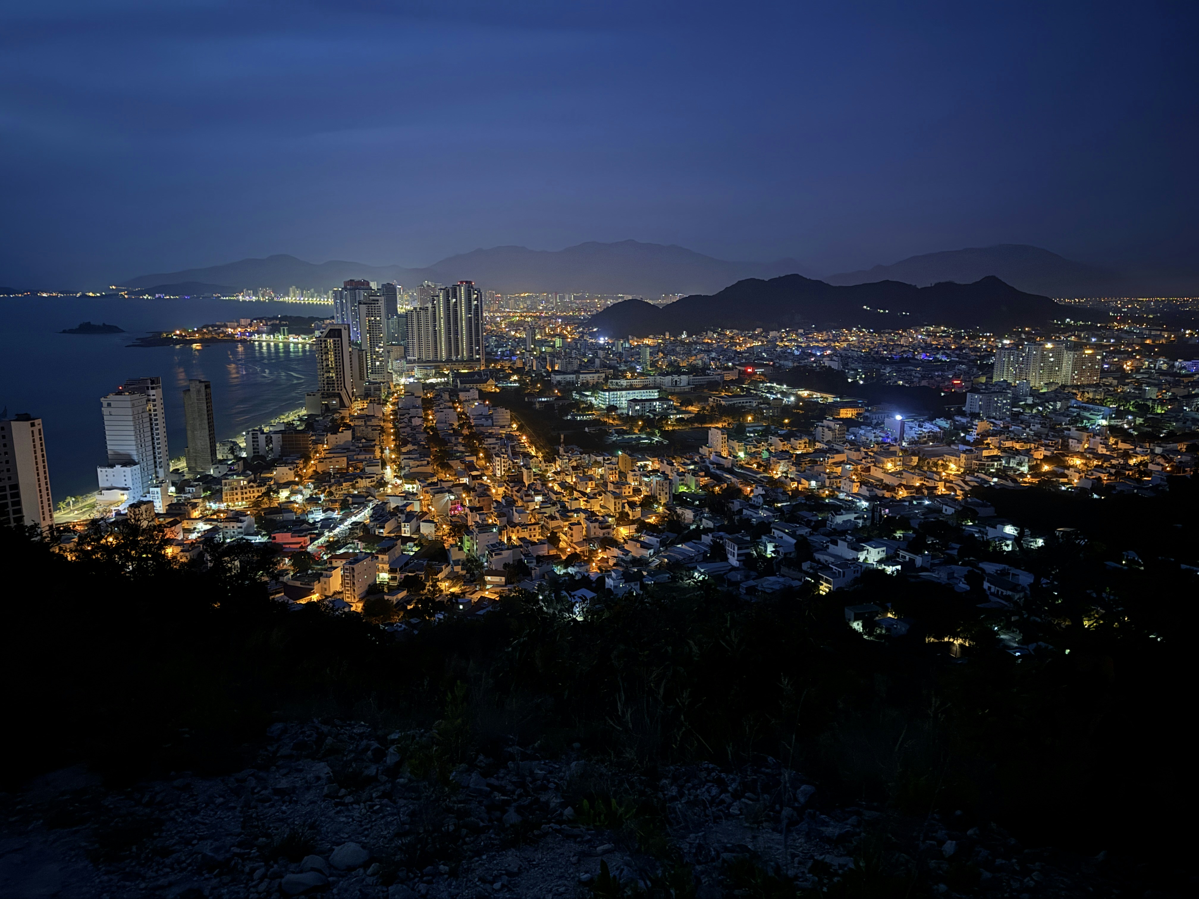 Cityscape at night with illuminated buildings and distant hills under a deep blue sky.