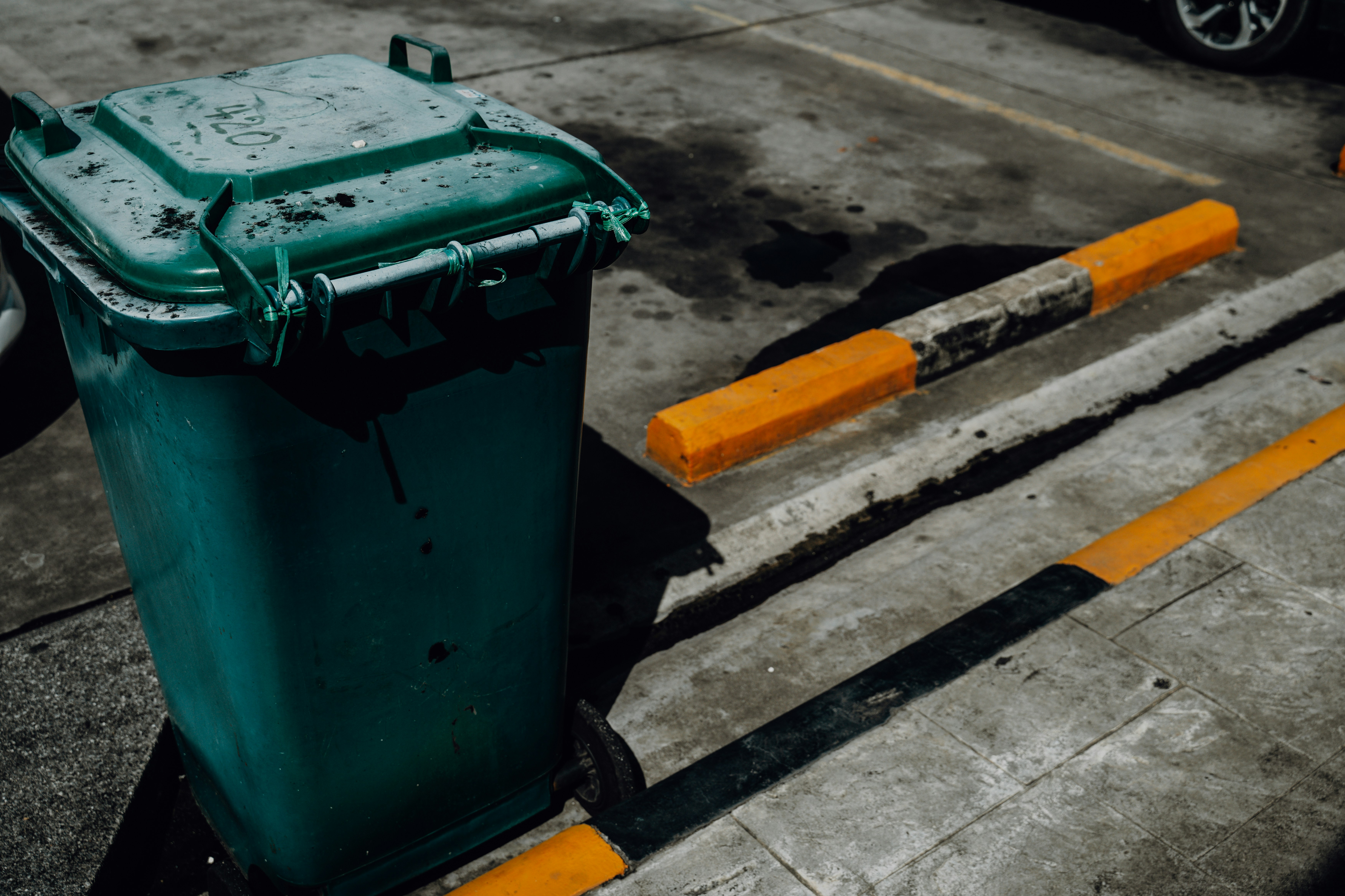 Green trash bin next to a yellow parking barrier casting shadows on a concrete surface.