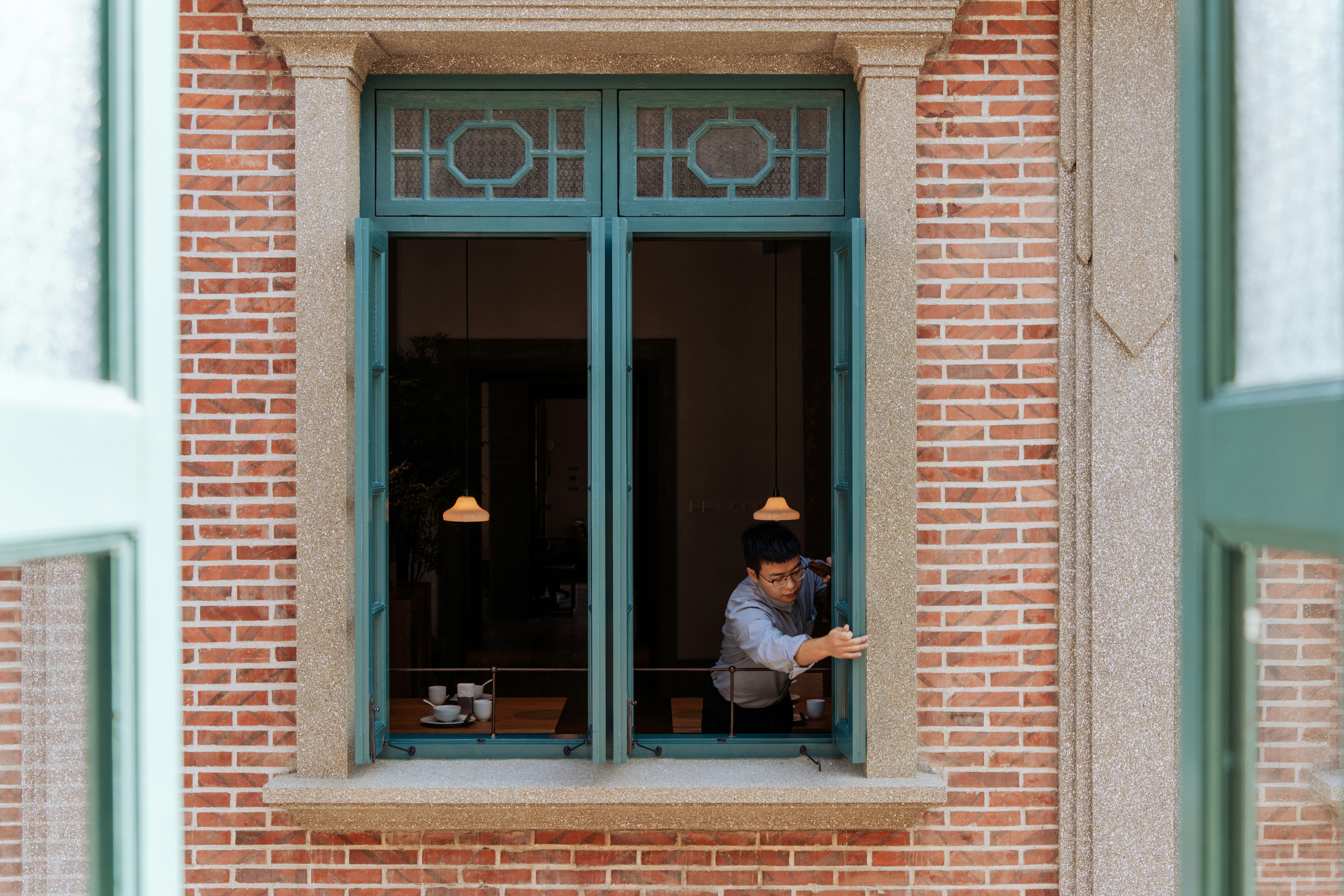 Person leaning out of an ornate window set in a brick wall, adjusting the window frame.