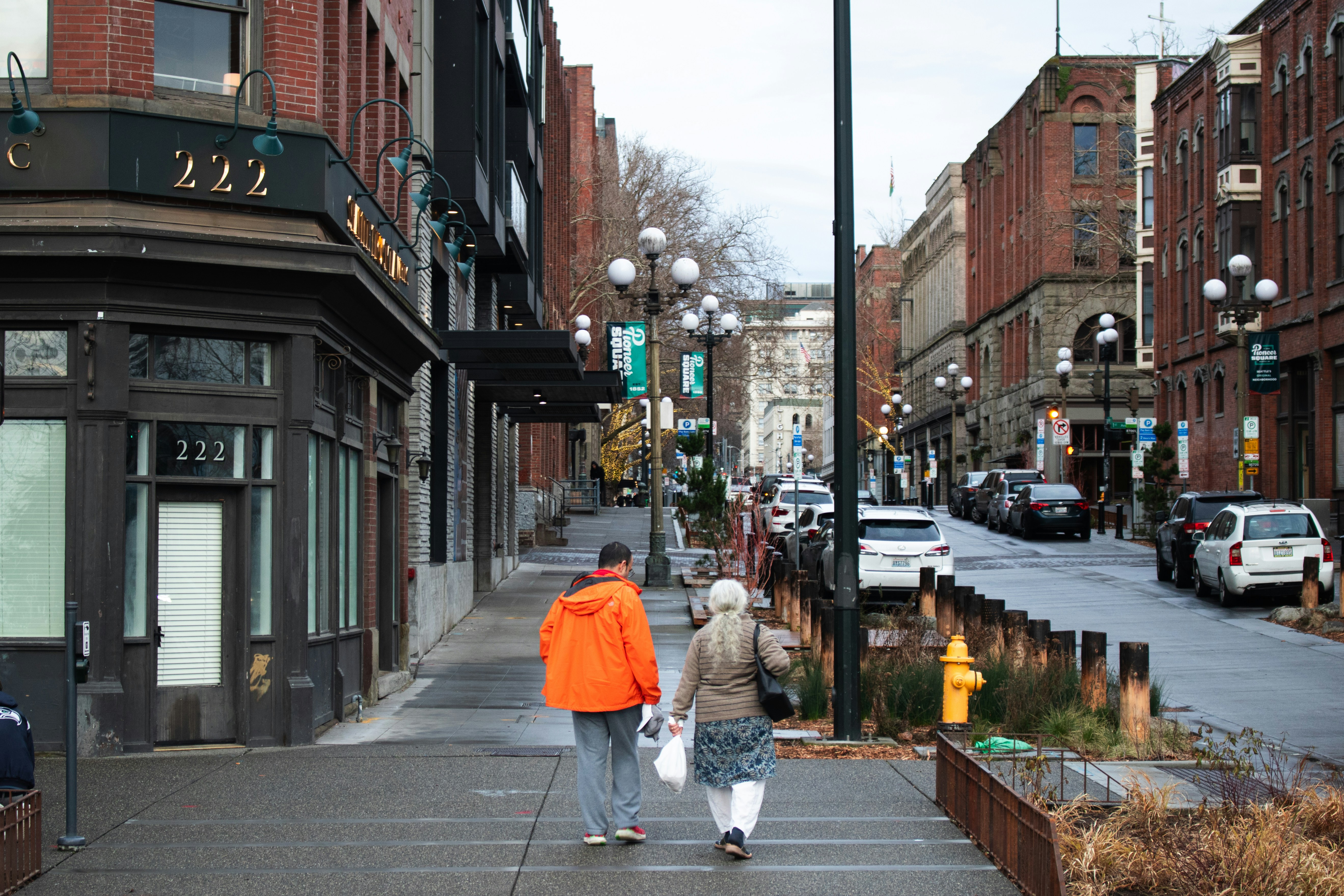 Two people walk along a wet city sidewalk lined with historic brick buildings and streetlamps.