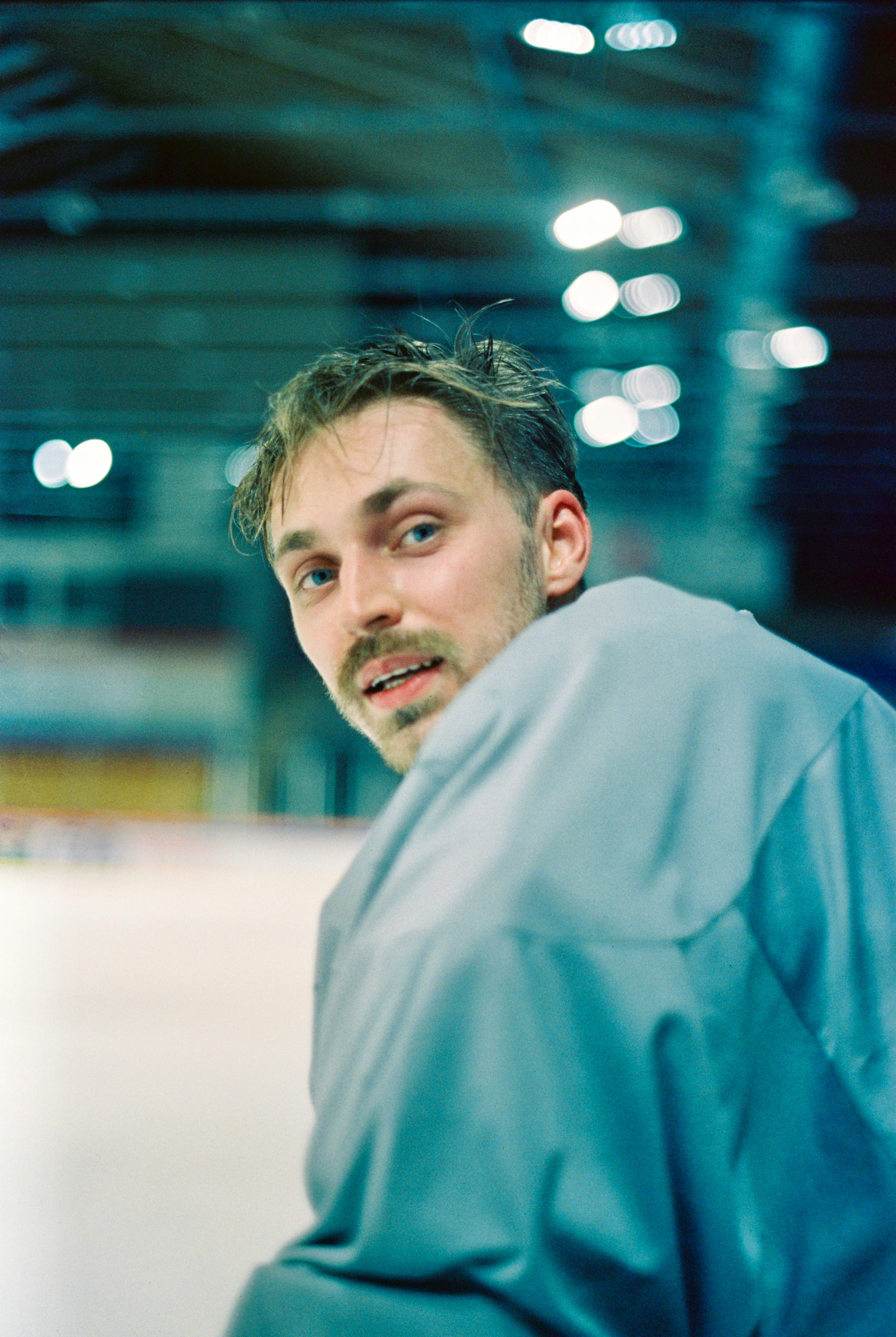Athlete in a gray jacket glancing back with a thoughtful expression, set against an ice rink backdrop. 