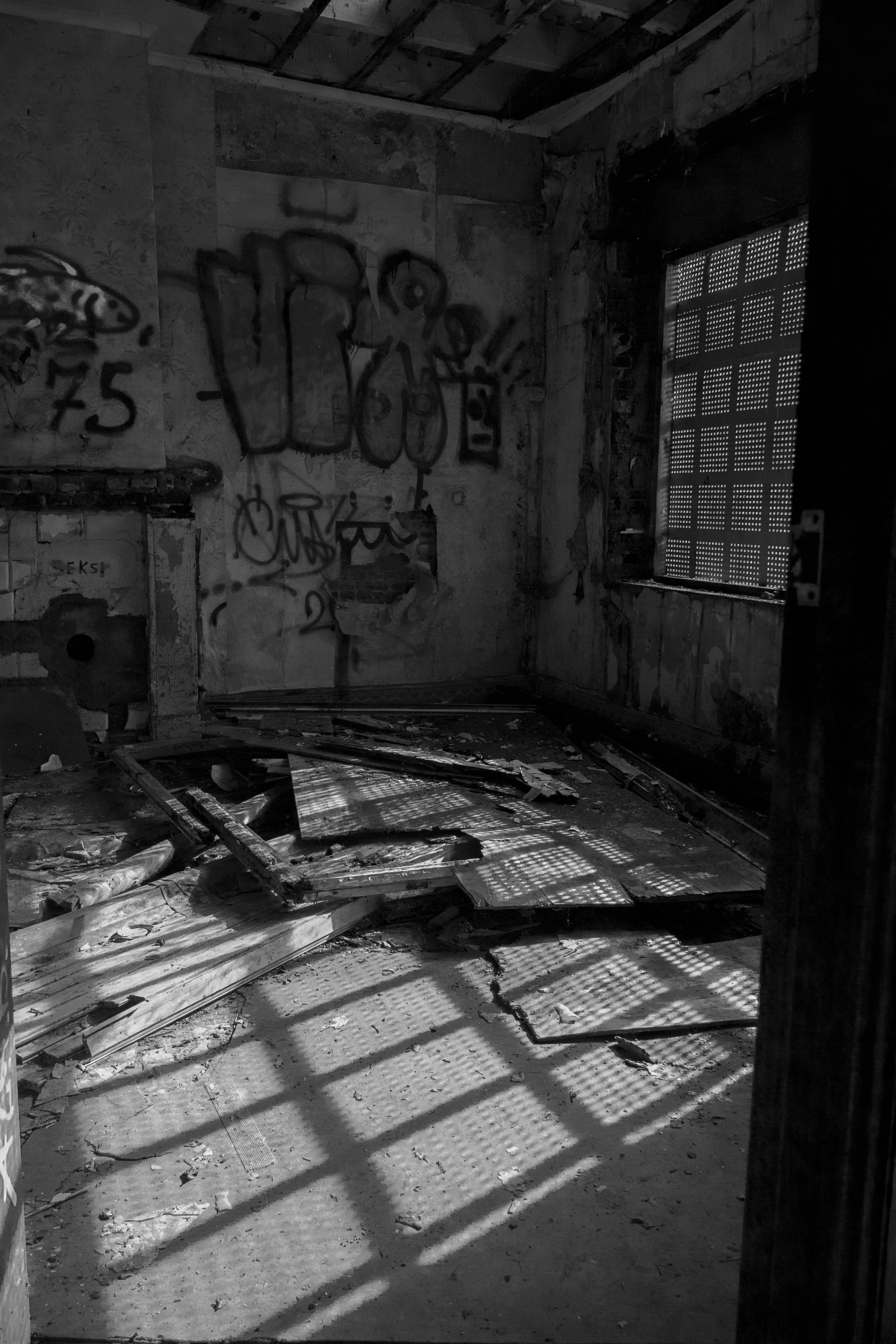 Dimly lit room with graffiti-covered walls and scattered debris, illuminated by patterned light streaming through a barred window.
