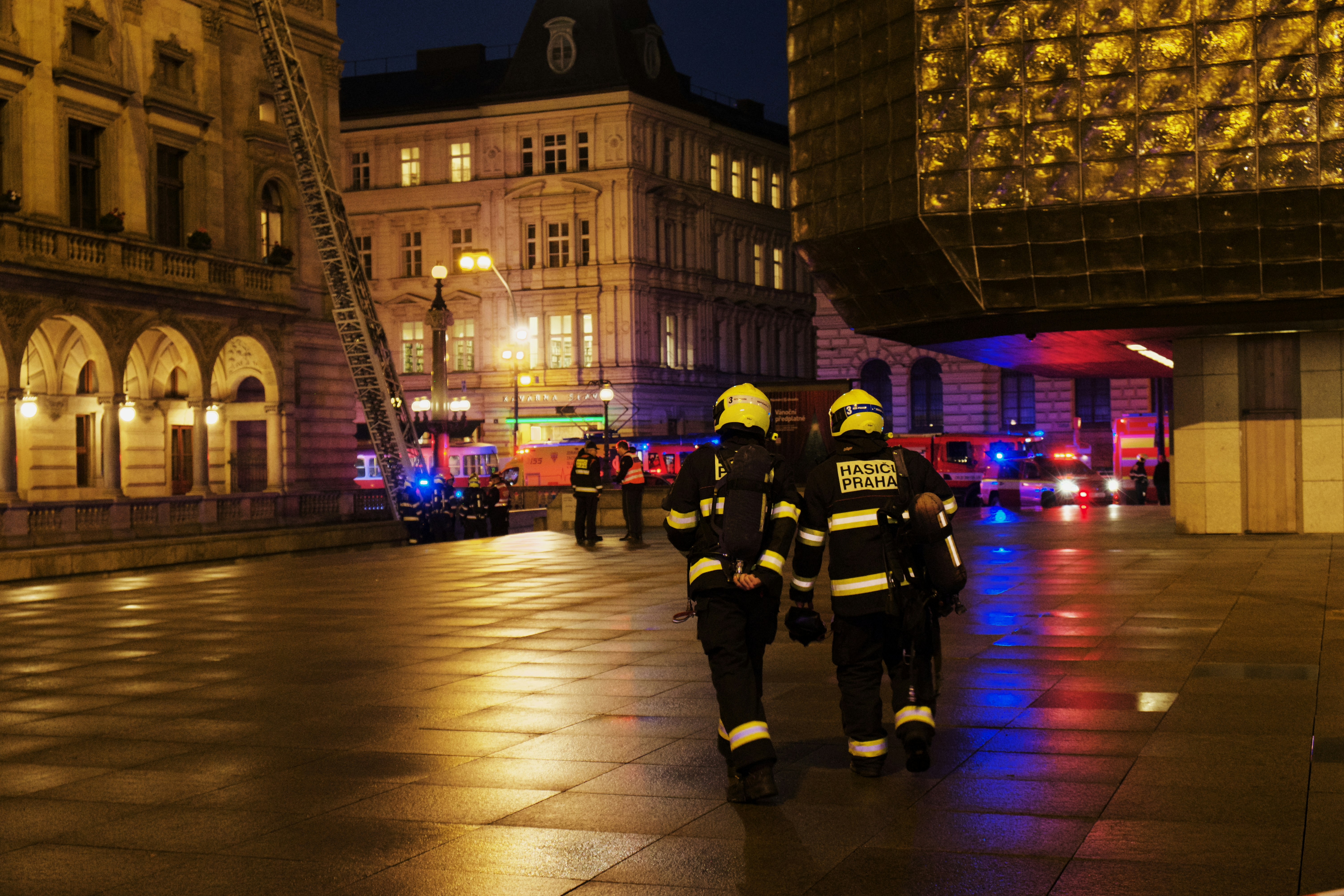 Firefighters walking through an urban square at night, with emergency vehicles illuminating the background.