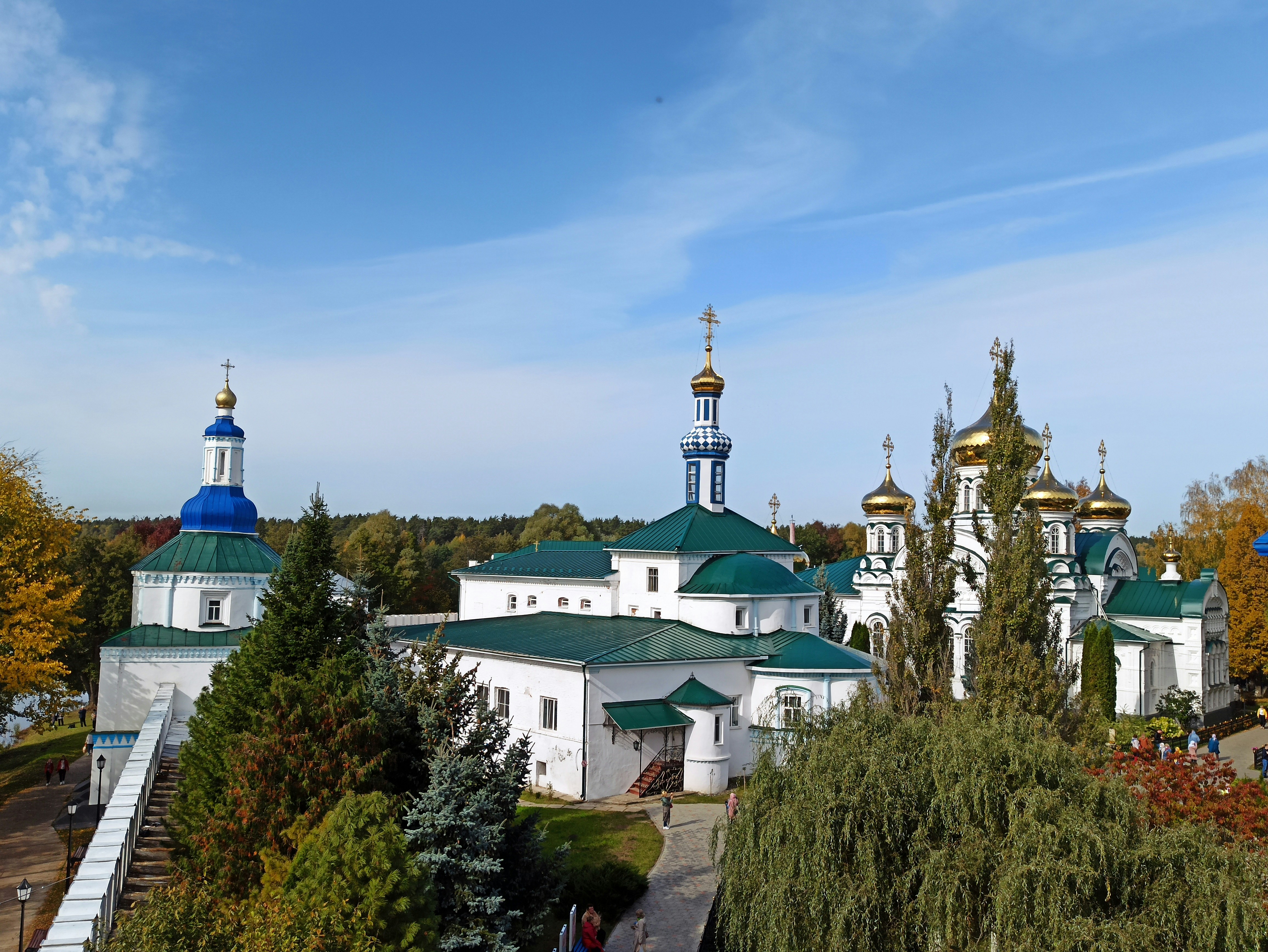 Orthodox church with green roofs and golden domes surrounded by autumn trees under a clear blue sky.