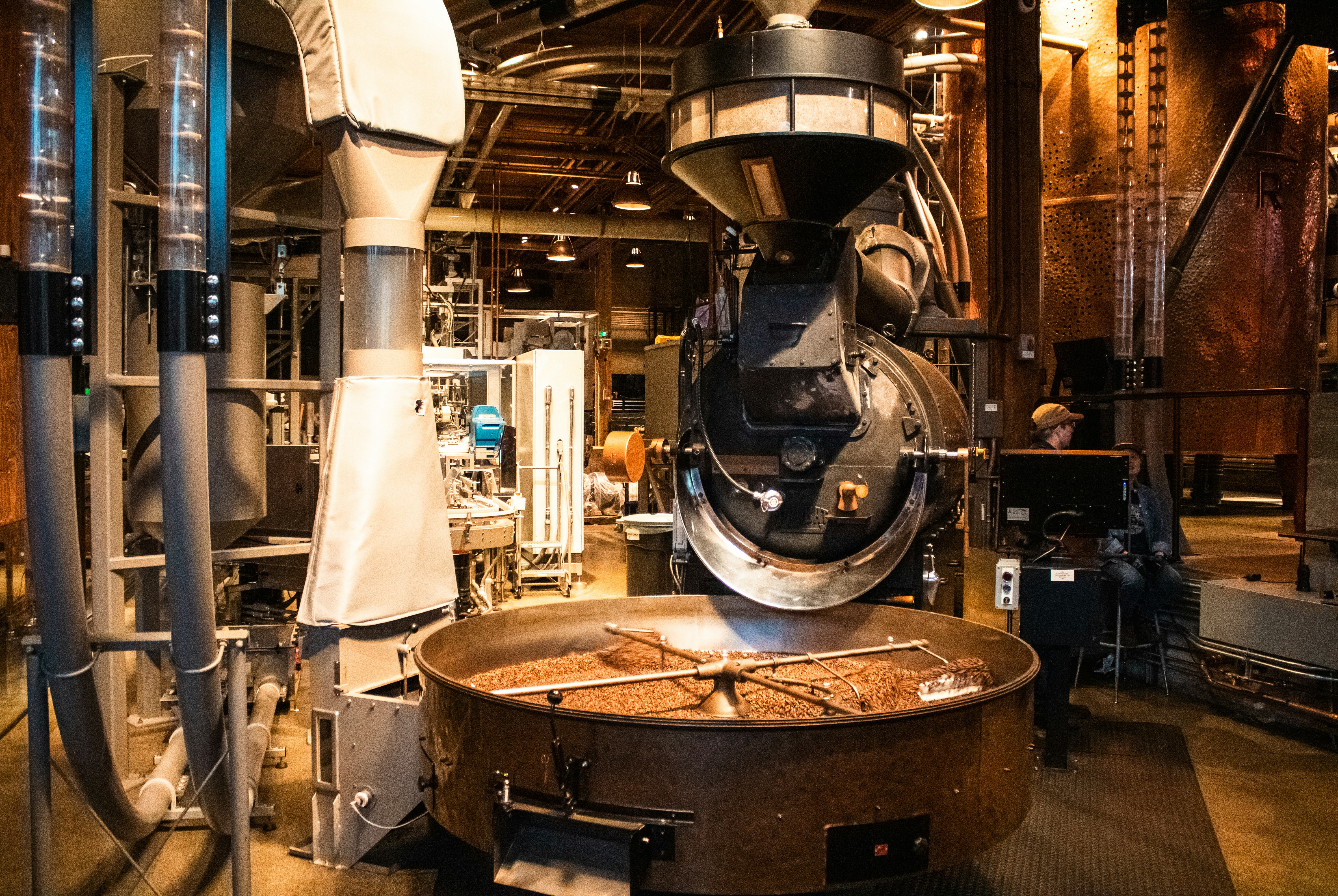 Industrial coffee roaster with freshly roasted beans in a cooling tray at a bustling roastery.