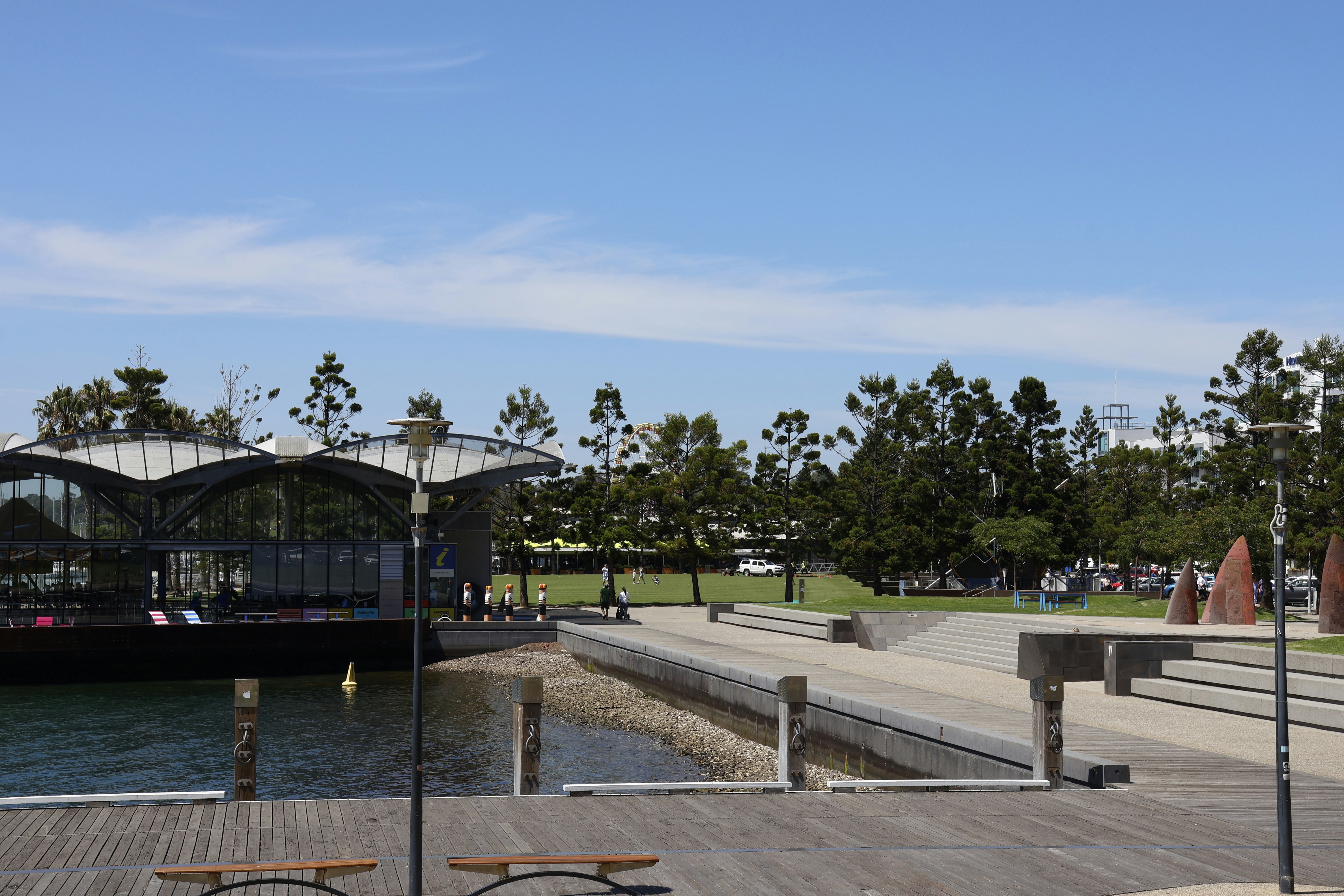 Contemporary pavilion structure beside a calm waterfront with lush greenery and clear blue sky.