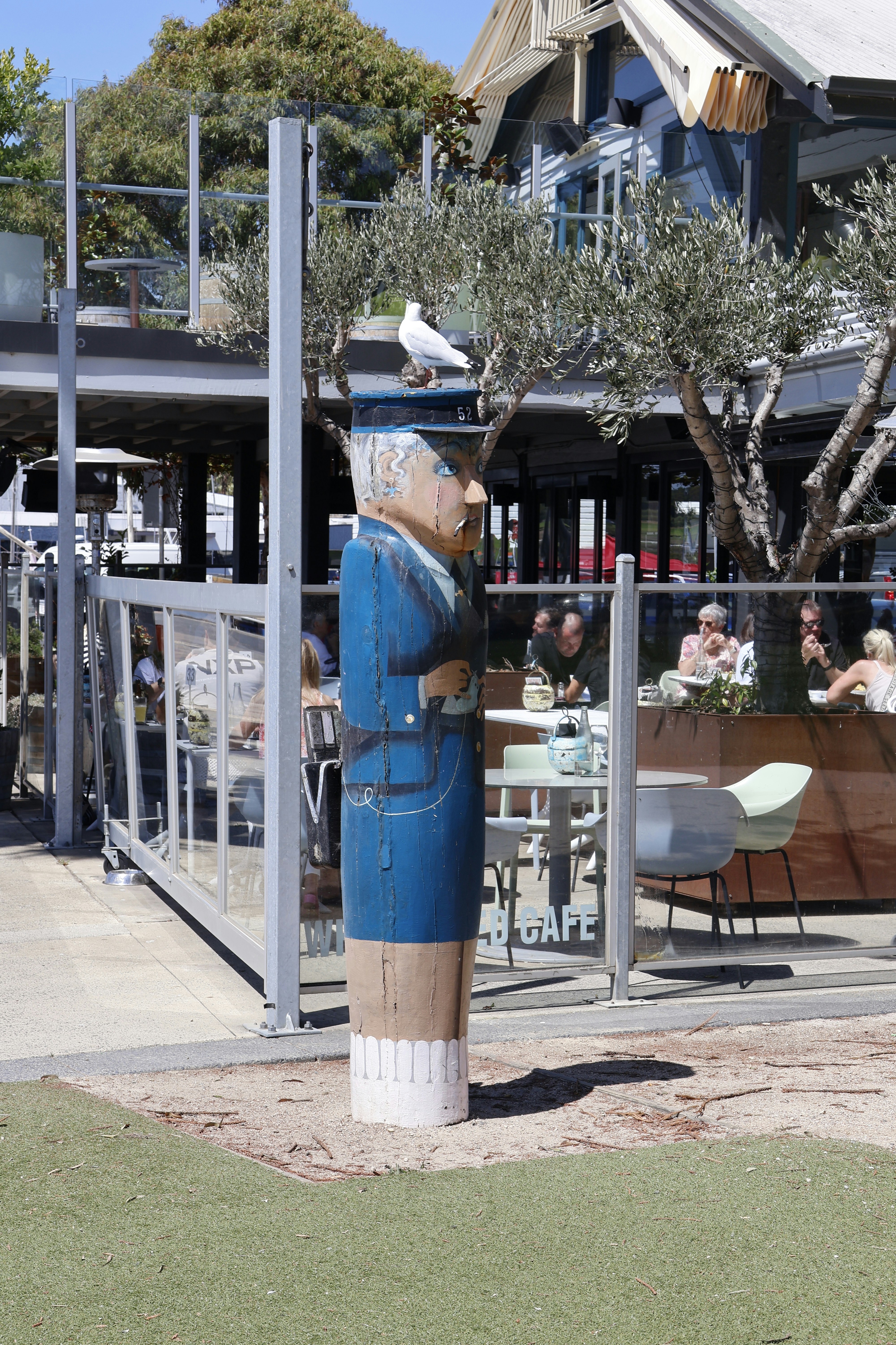 Wooden sculpture of a uniformed figure with a seagull perched on its hat near an outdoor cafe.