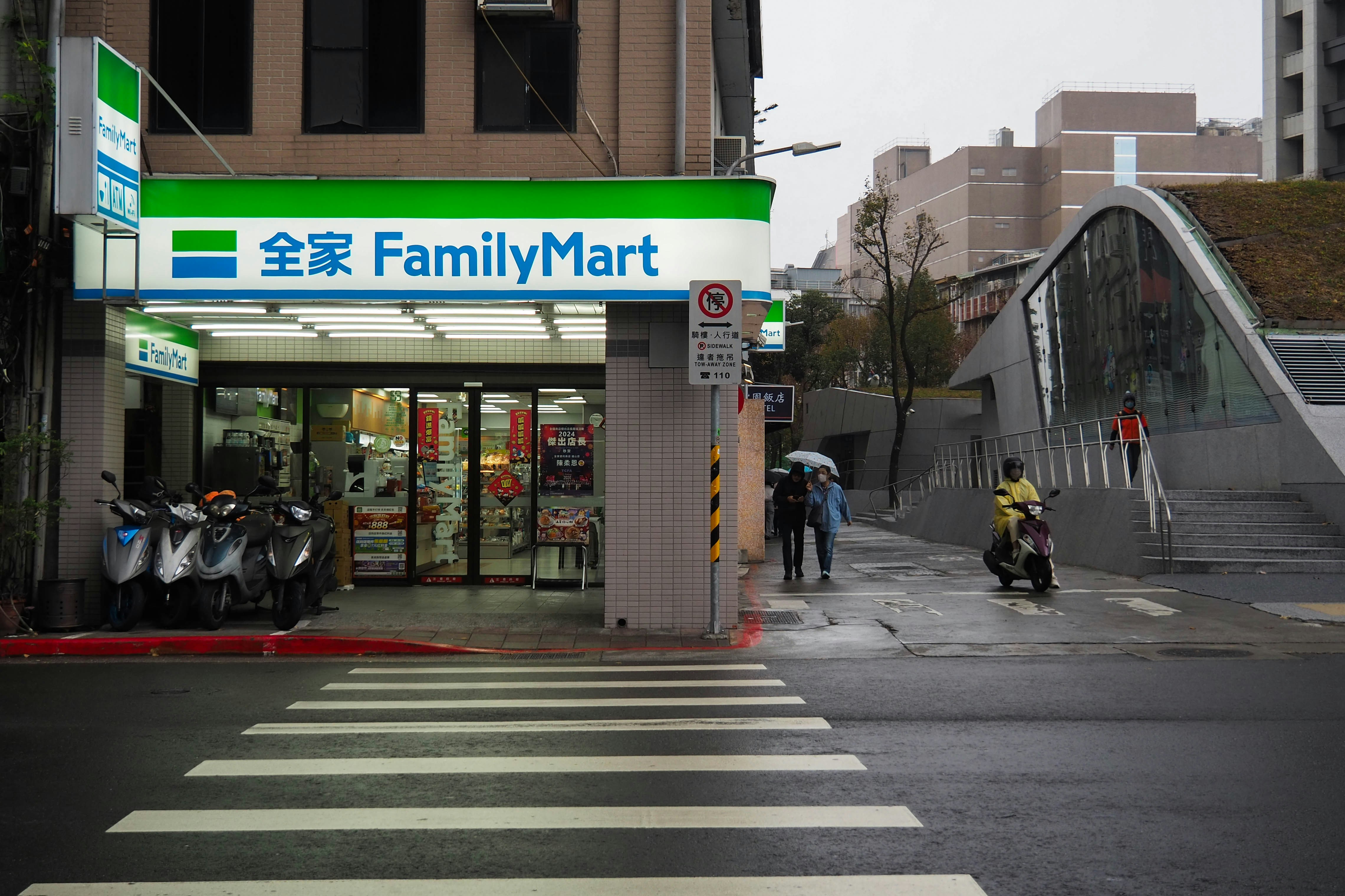 FamilyMart storefront on a rainy street with a motorcyclist passing by and an arched staircase in the background.
