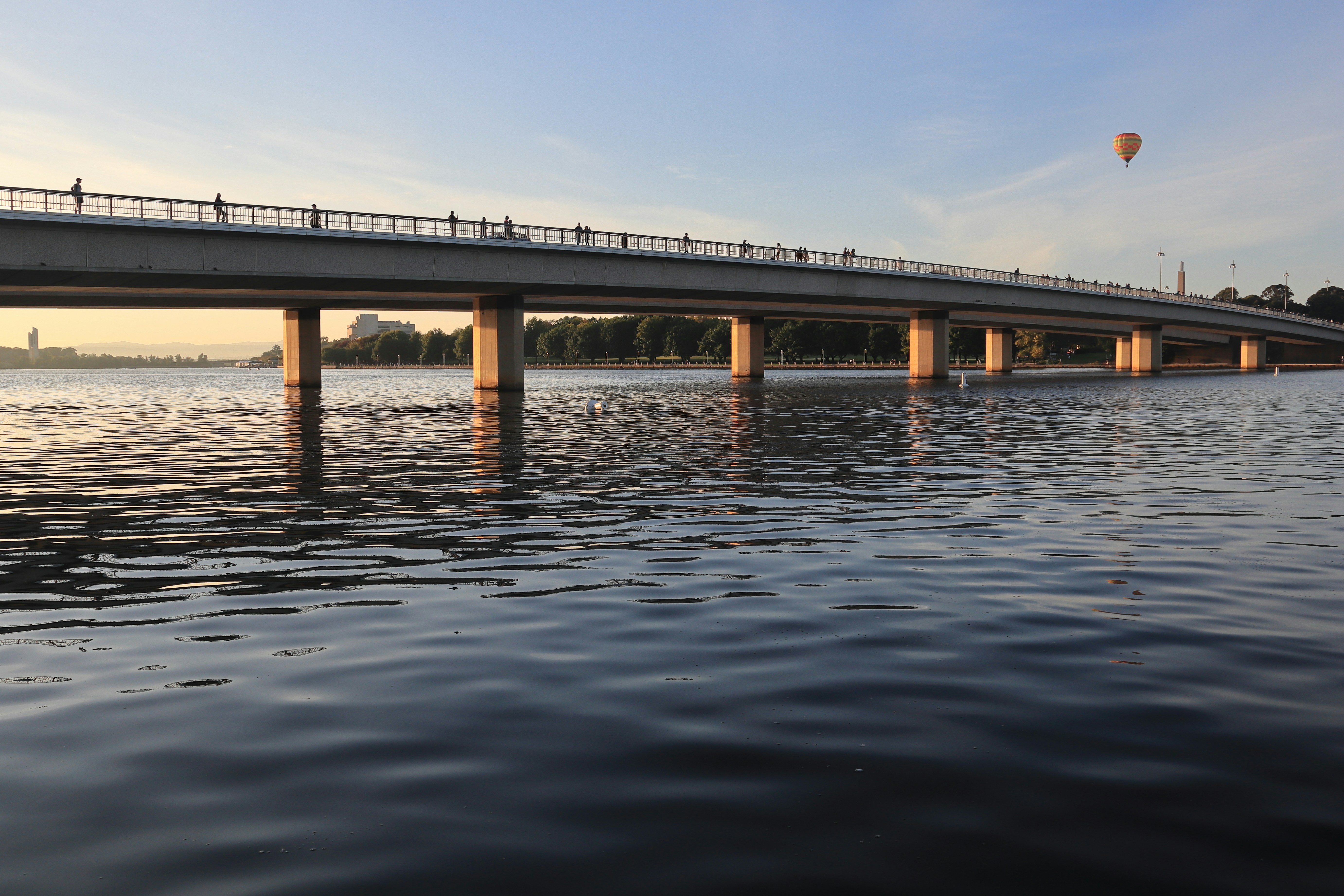 Bridge spanning calm water with a hot air balloon in the sky at sunset.