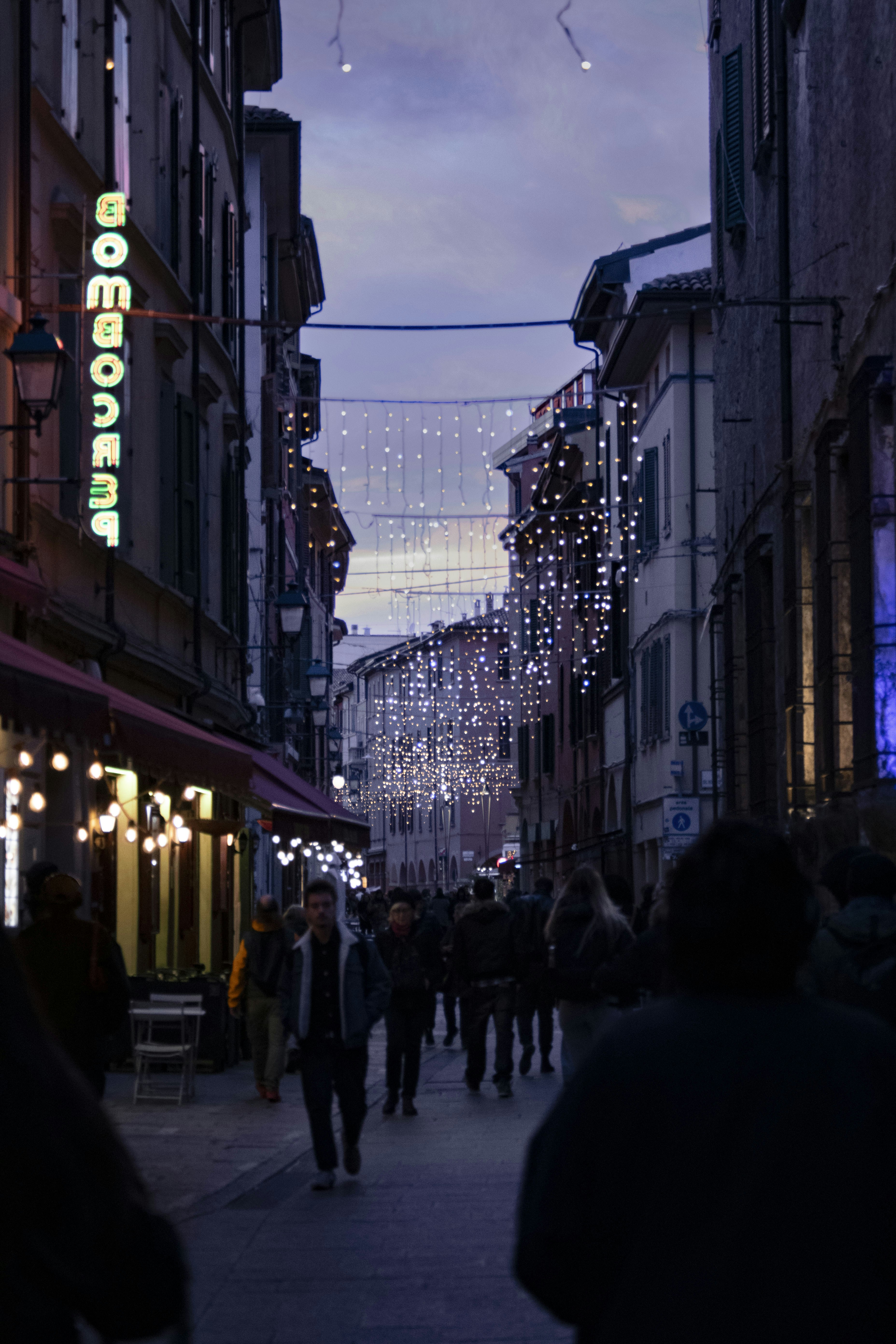Pedestrians walking under twinkling string lights in a narrow street at dusk.