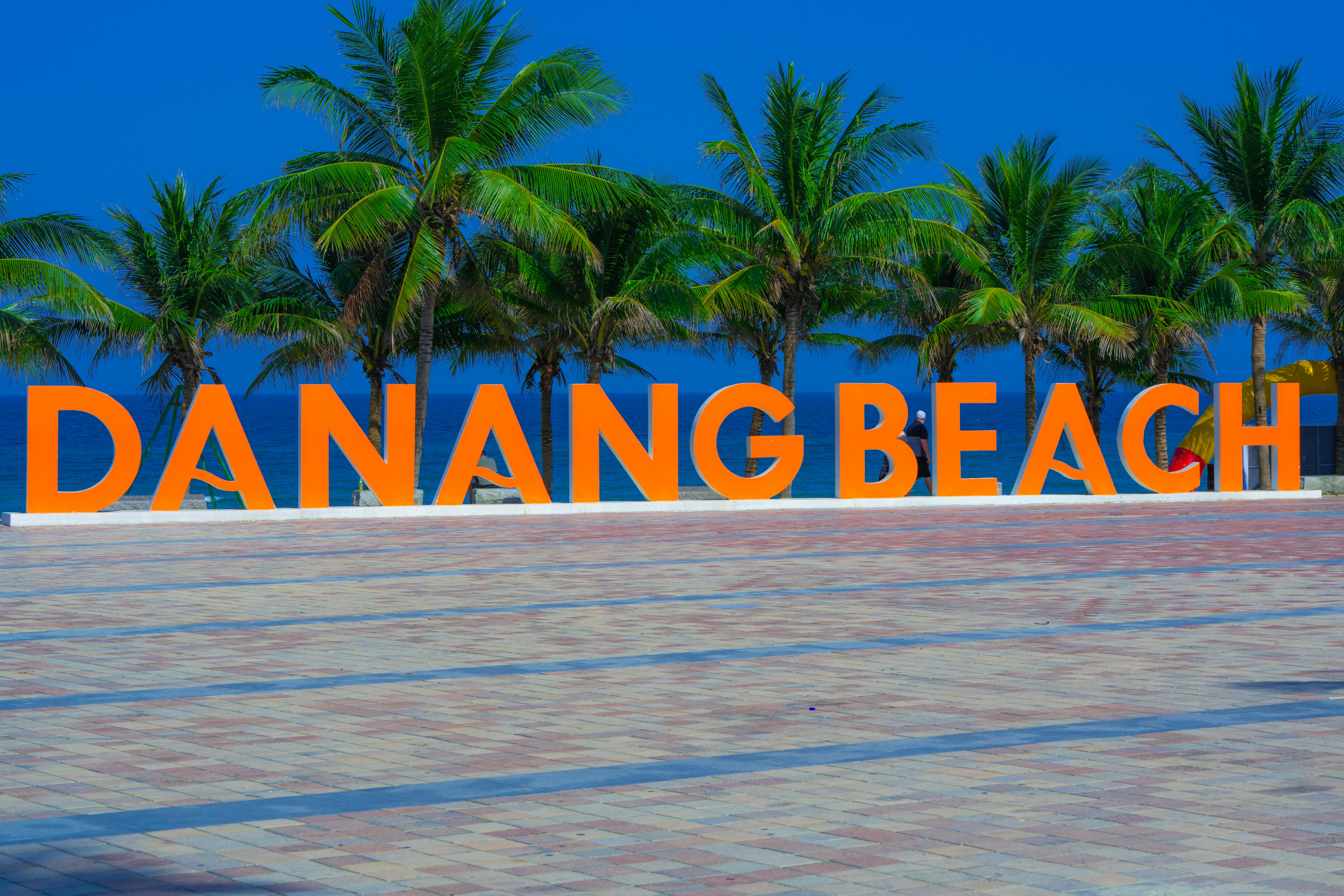 Bold orange Danang Beach sign framed by lush palm trees against a vivid blue sky.