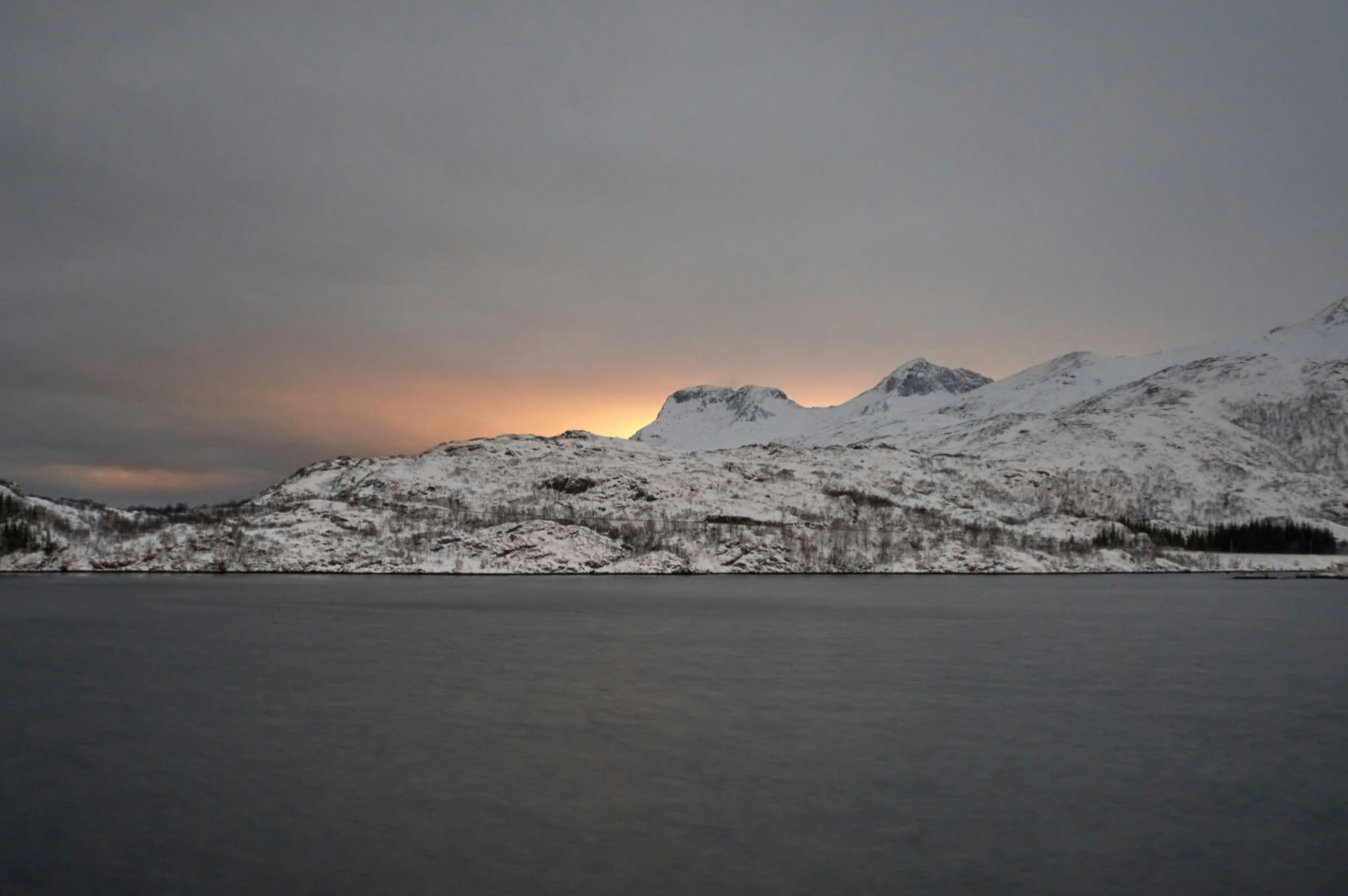 Snow-covered mountains by the sea illuminated by a soft sunset glow.
