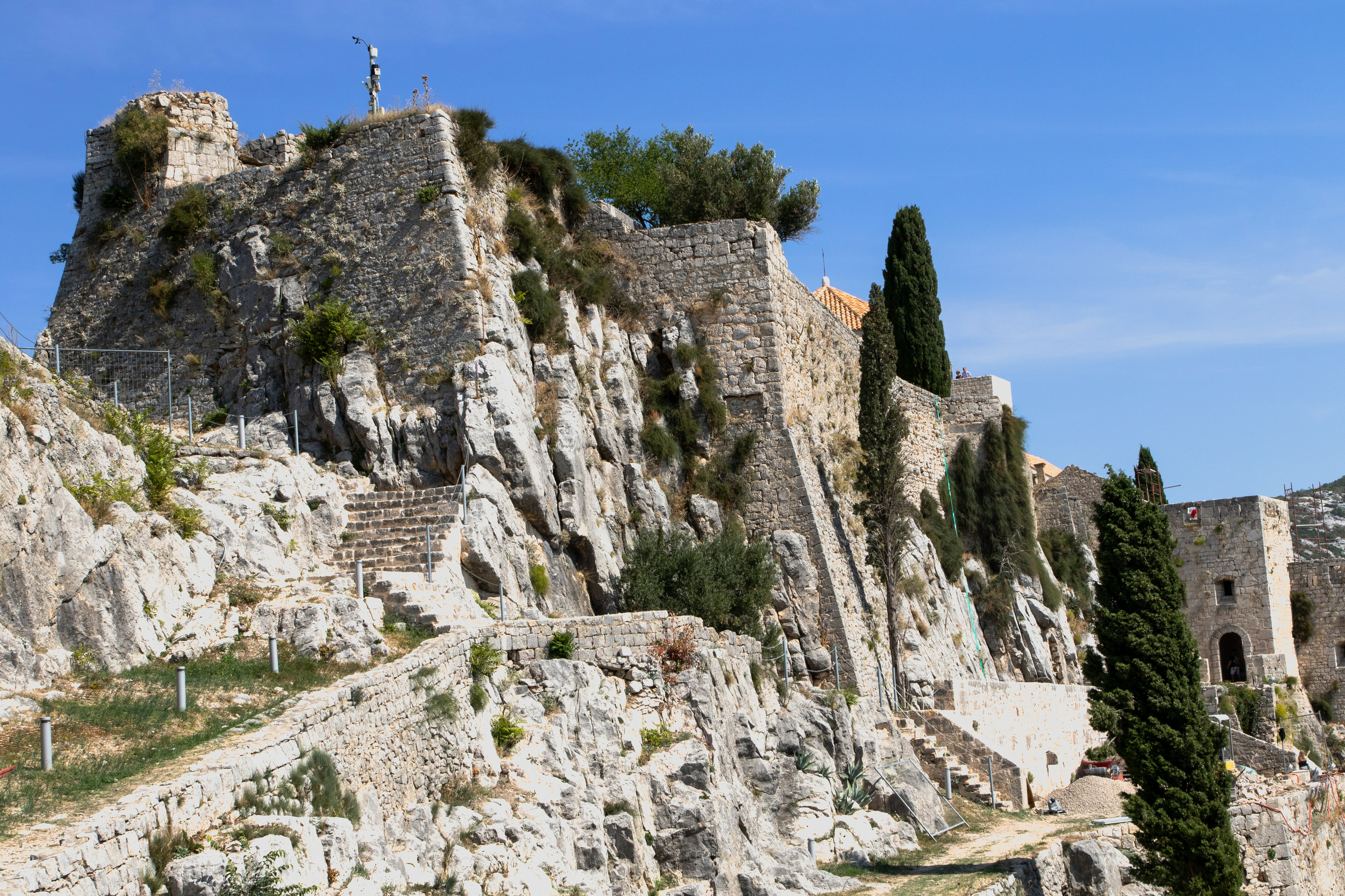 Stone fortress perched on rocky cliffs with lush greenery and winding steps leading to its entrance.