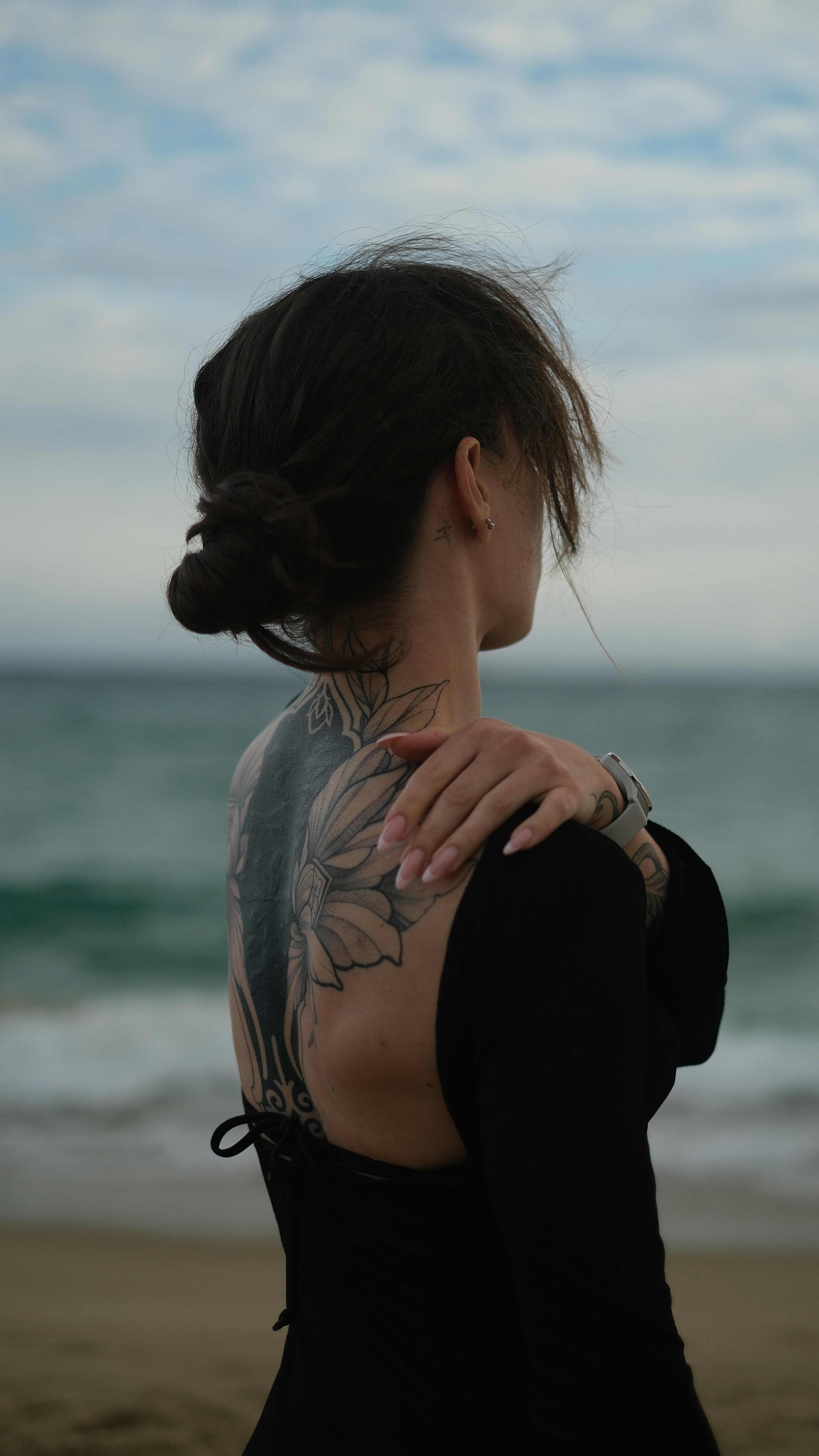 Portrait of a tattooed woman seen from behind on a sandy beach with waves in the background. The shot emphasizes the floral-back tattoo and the subject's calm, poised demeanor.
