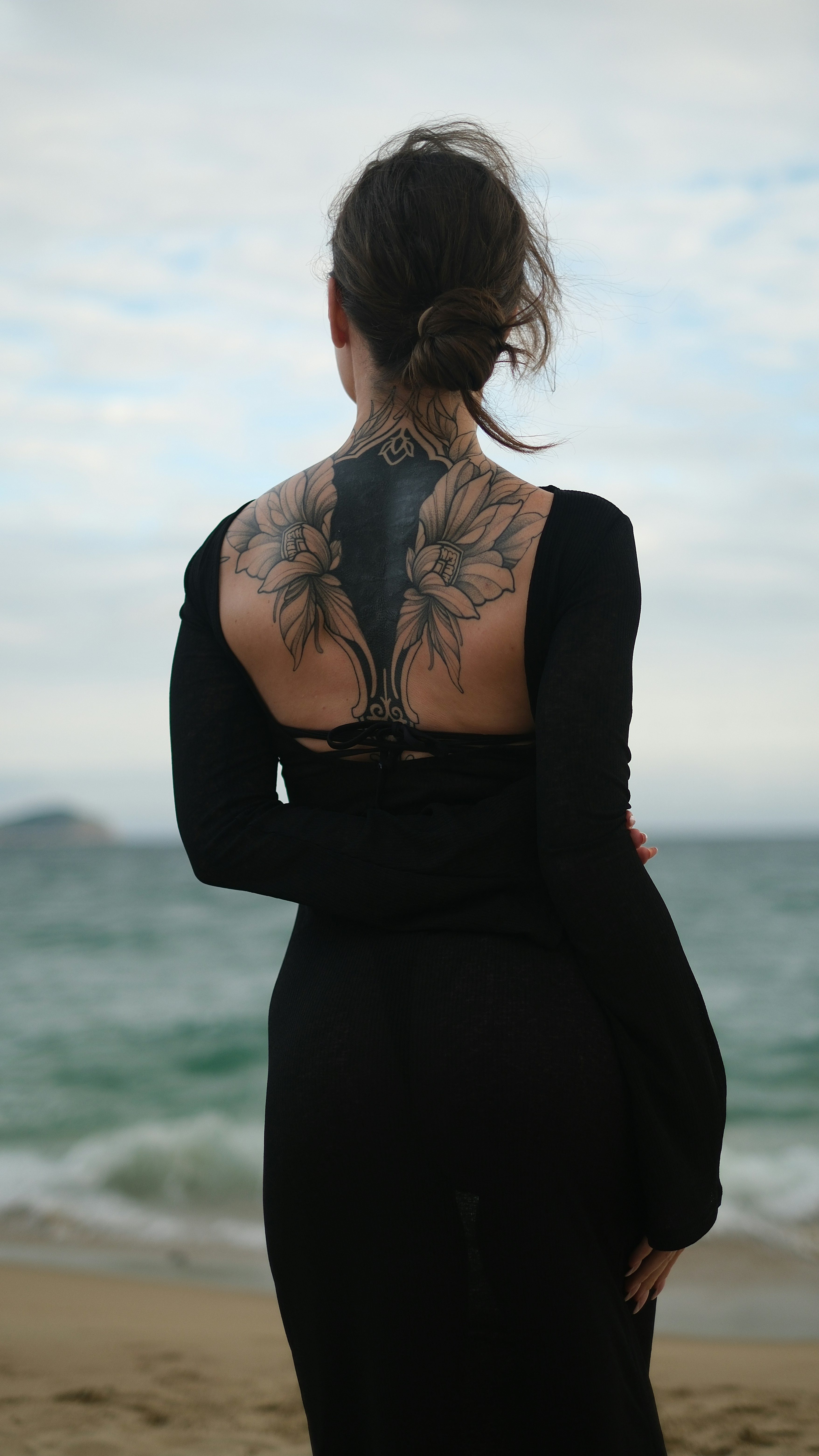 Photograph of a person with a large floral tattoo on the back, standing on a sandy beach, facing the ocean in a black outfit.