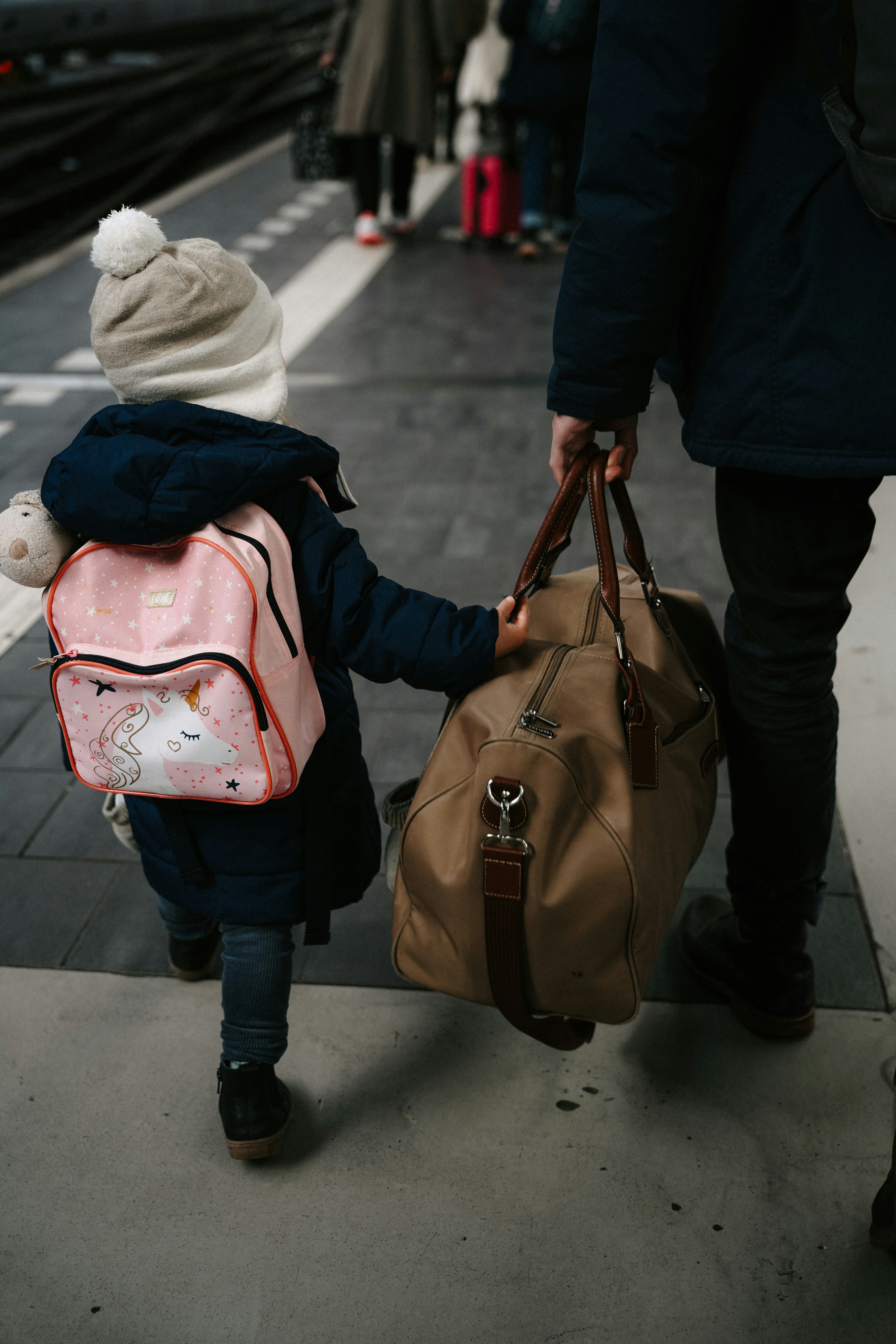 Child wearing a pink backpack holds an adult's hand while walking on a train platform.