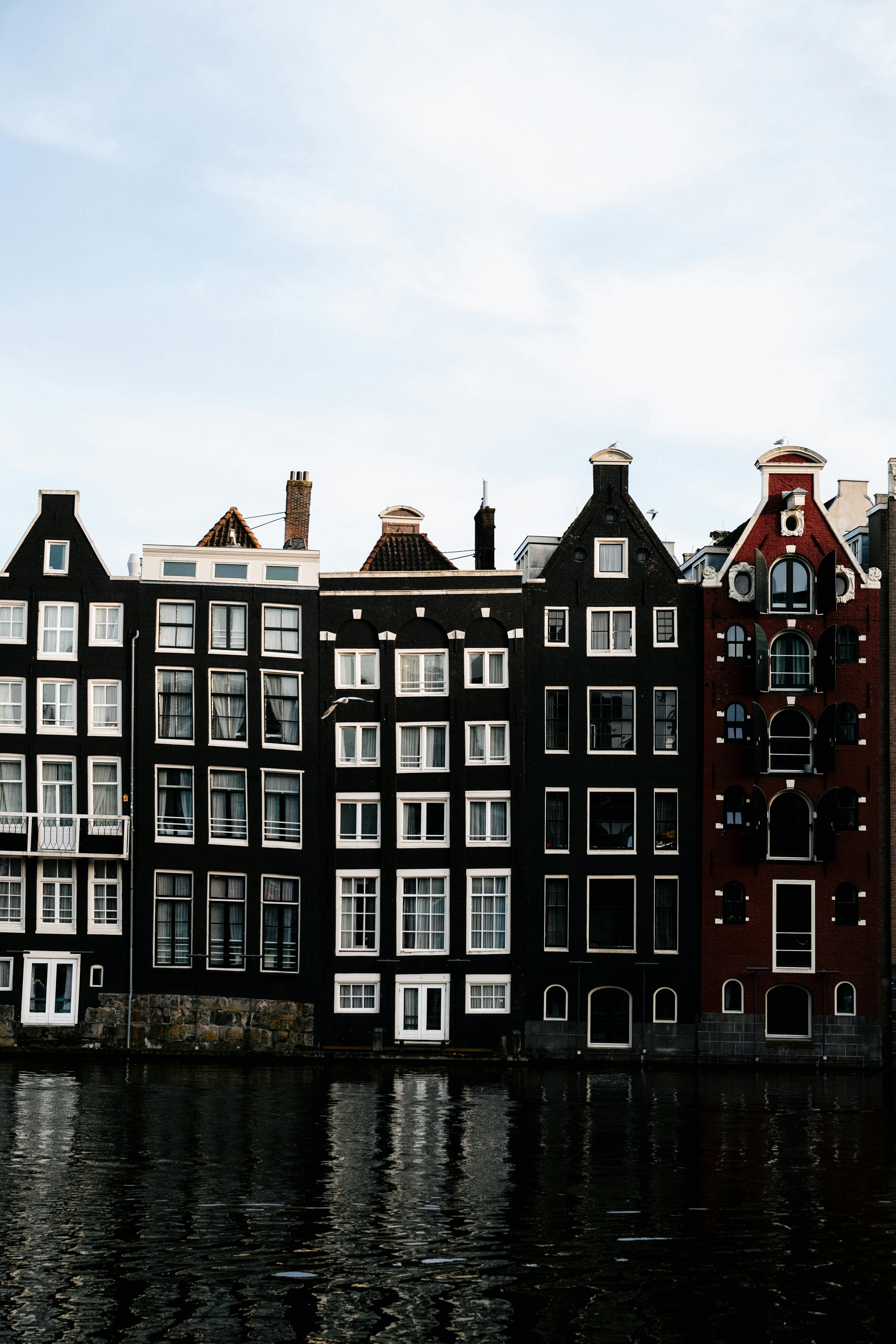 Historic canal-side buildings with distinctive gabled roofs reflecting in calm water under a bright sky.