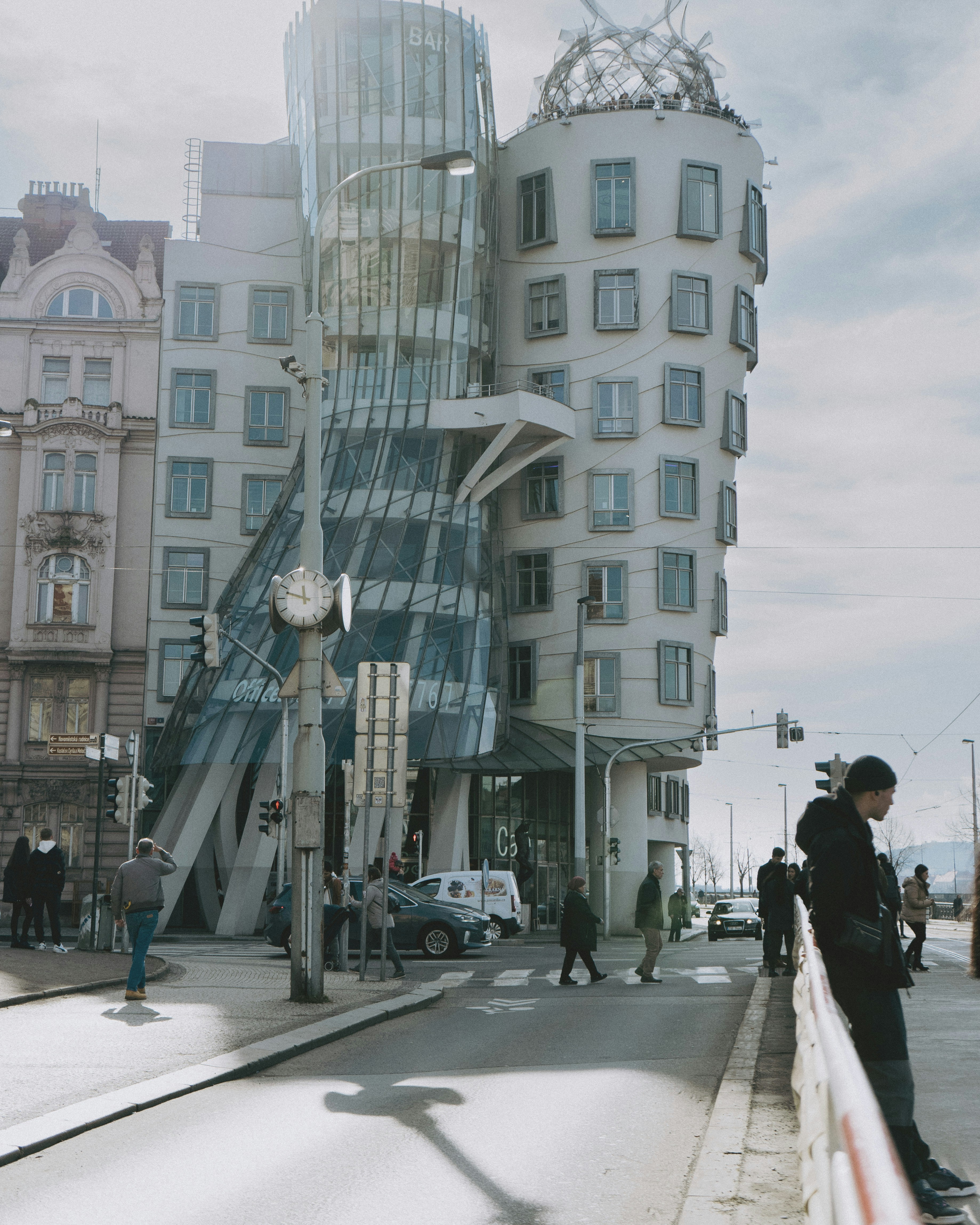 Modern, uniquely curved building contrasts with classical architecture under a cloudy sky.