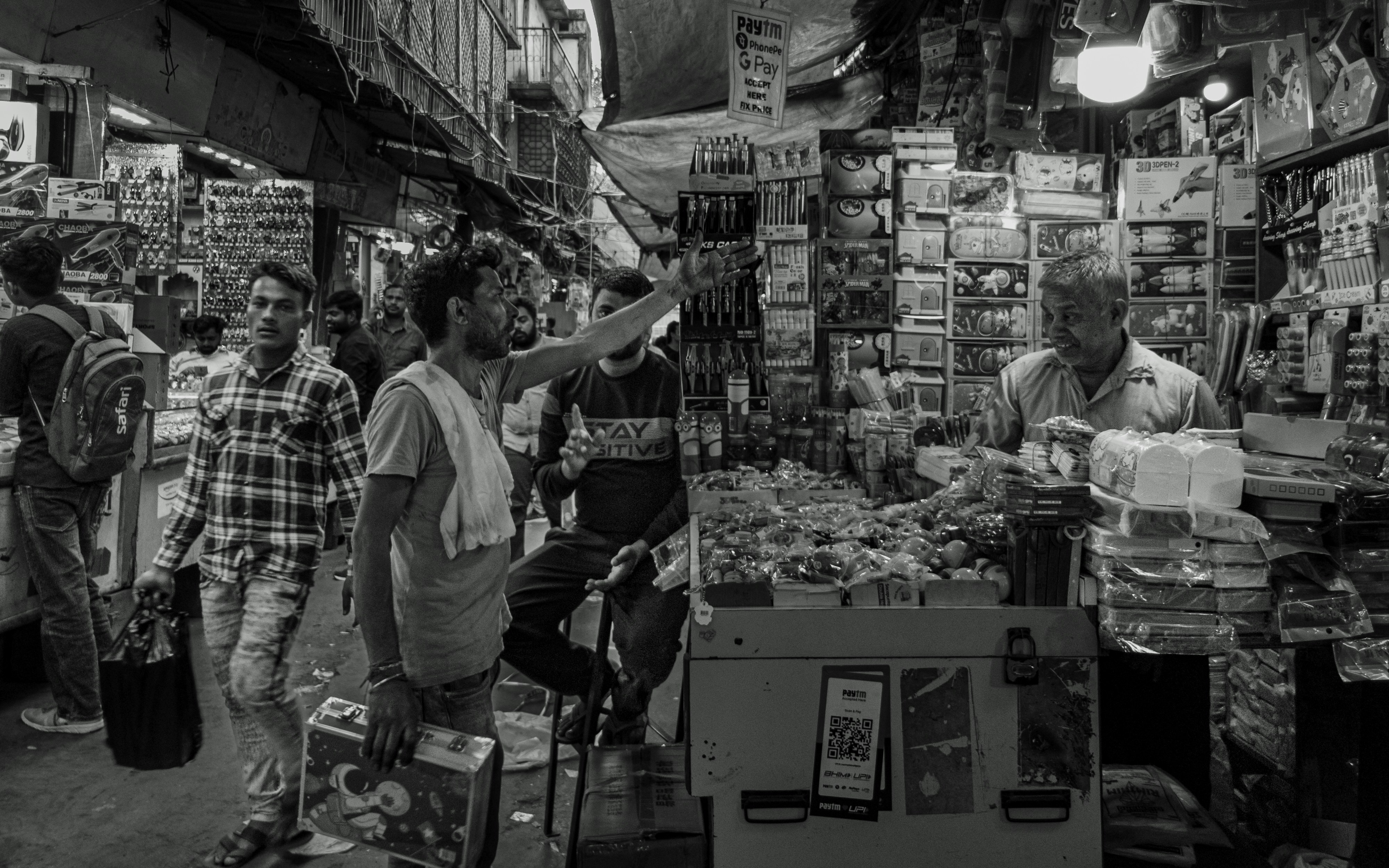 Bustling market scene with vendors and shoppers surrounded by stacks of magazines and goods under warm lighting.
