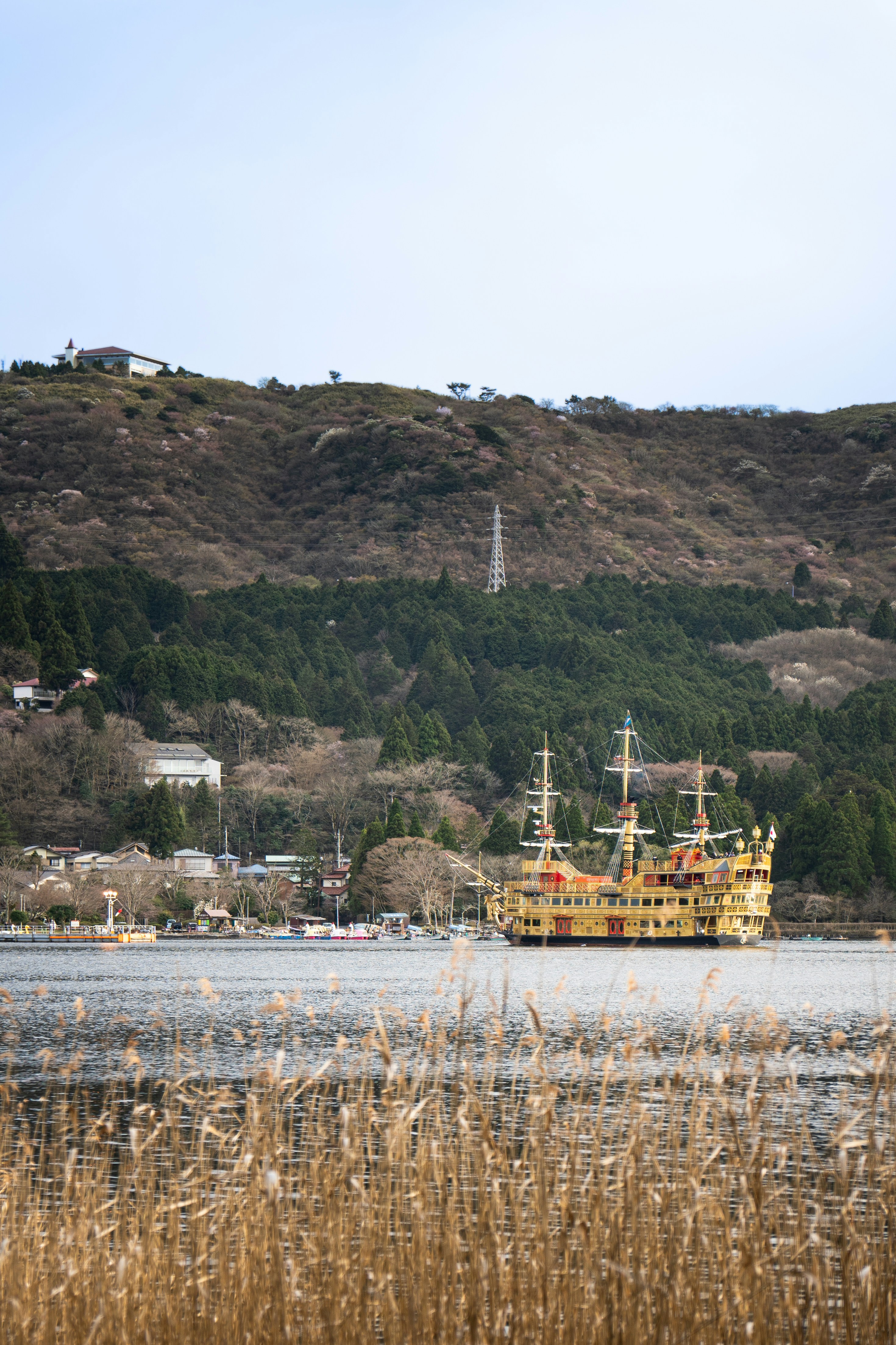A scenic view of a golden pirate ship sailing on Lake Ashi, surrounded by lush mountains and a peaceful lakeside village in Hakone, Japan.