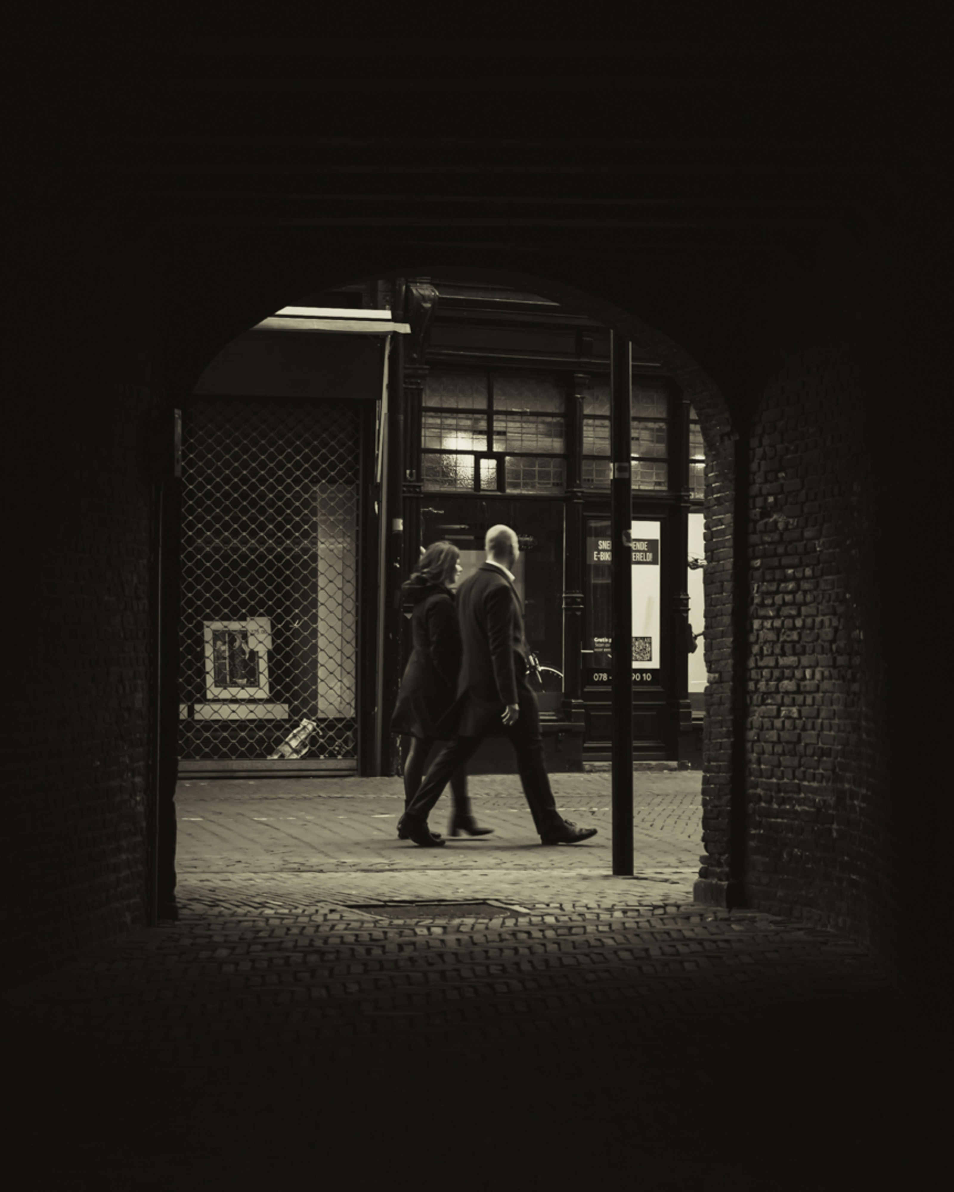 Beneath the Brick Arch, Two Figures Step into a Quiet City Street
