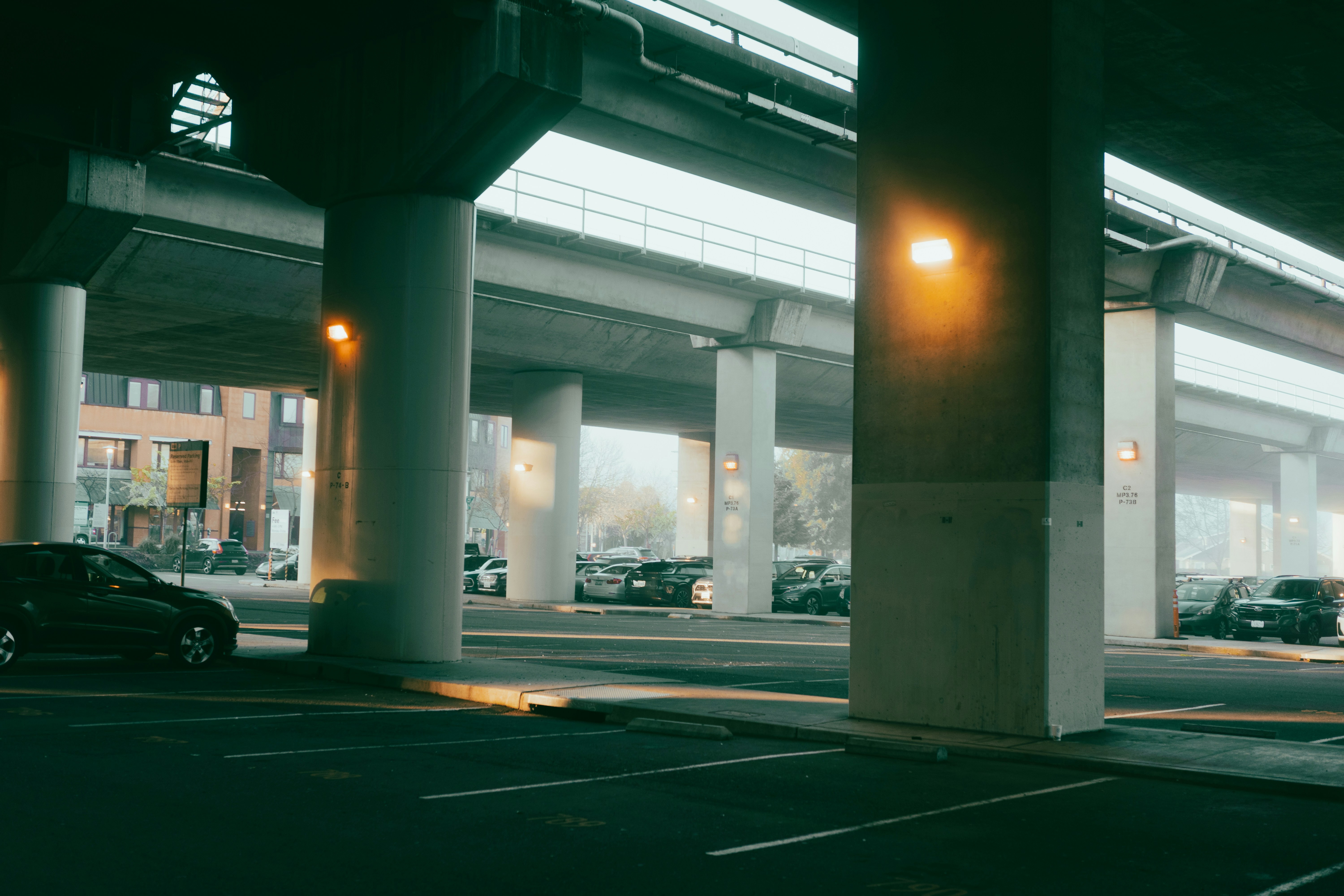 Dimly lit parking area beneath an overpass with structural columns and parked cars.