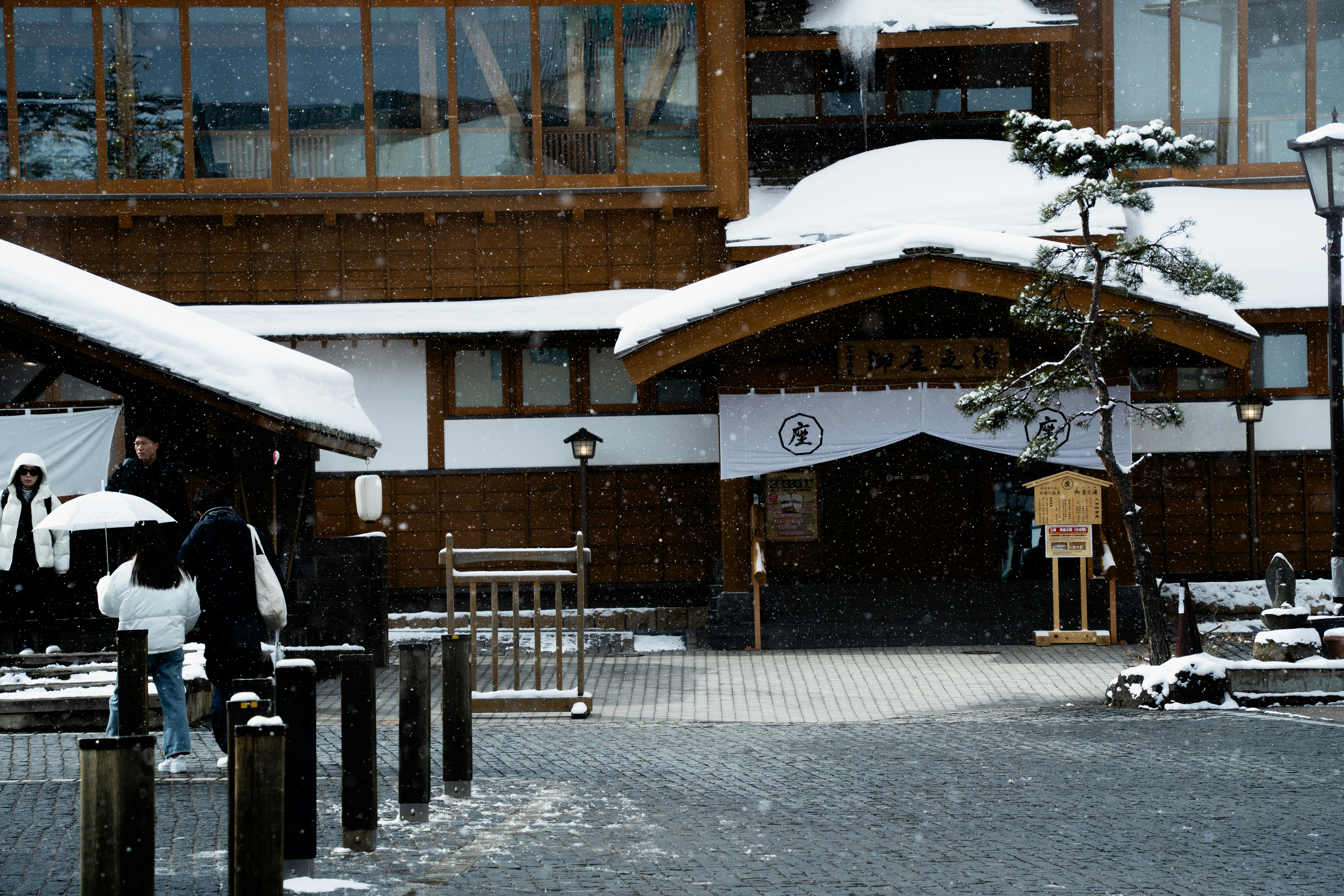Snowfall envelops a wooden building with a canopy and pine tree, as people walk with umbrellas.