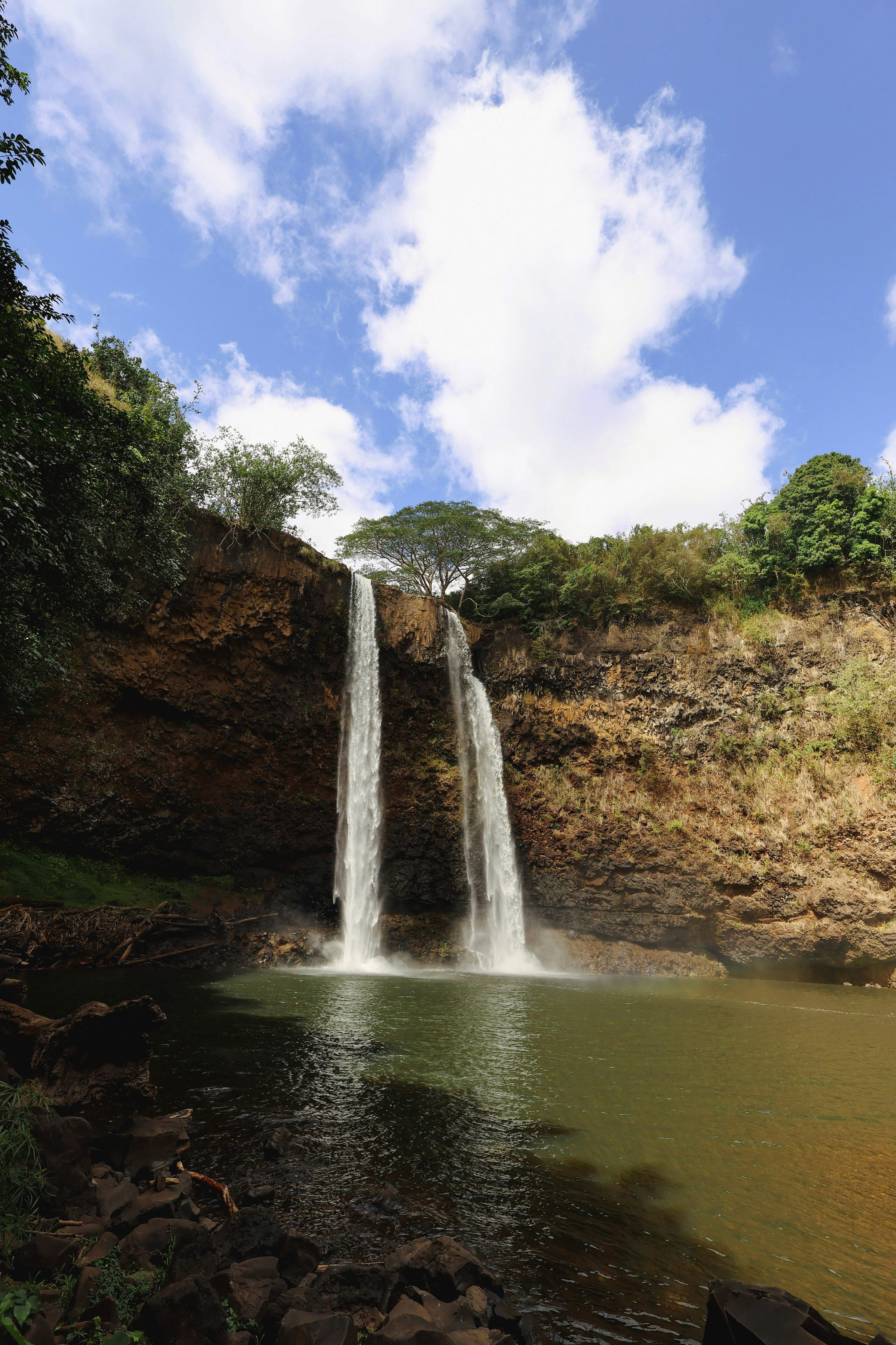 Twin waterfalls cascade into a tranquil pool surrounded by lush greenery under a bright blue sky.