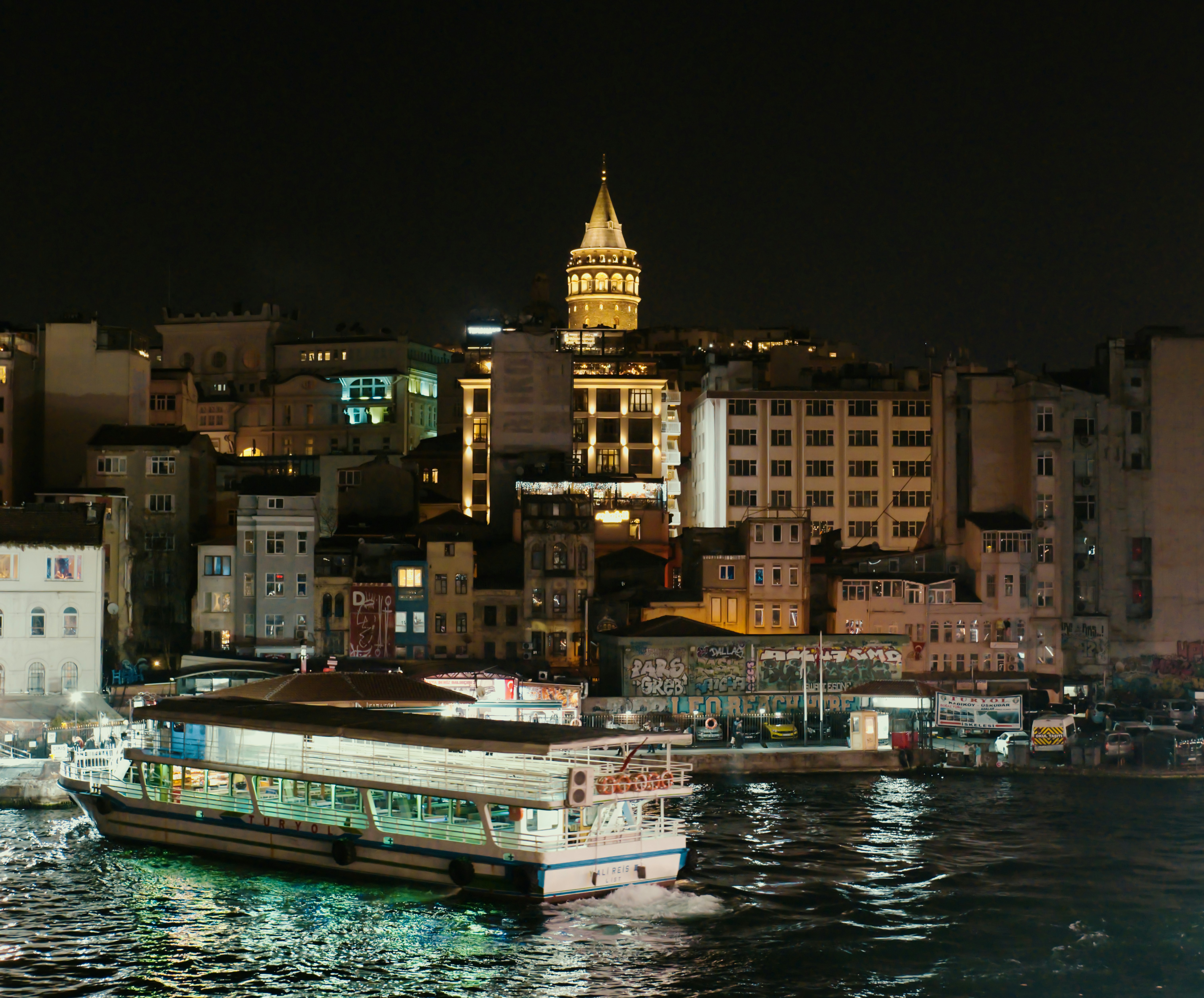 Nighttime cityscape featuring illuminated buildings and a prominent tower reflected in the water.