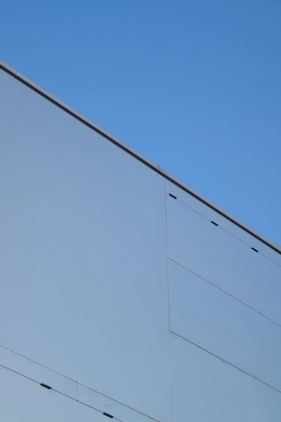 Diagonal white building facade with thin seam lines set against a clear blue sky. Small dark fixtures punctuate the upper edge.